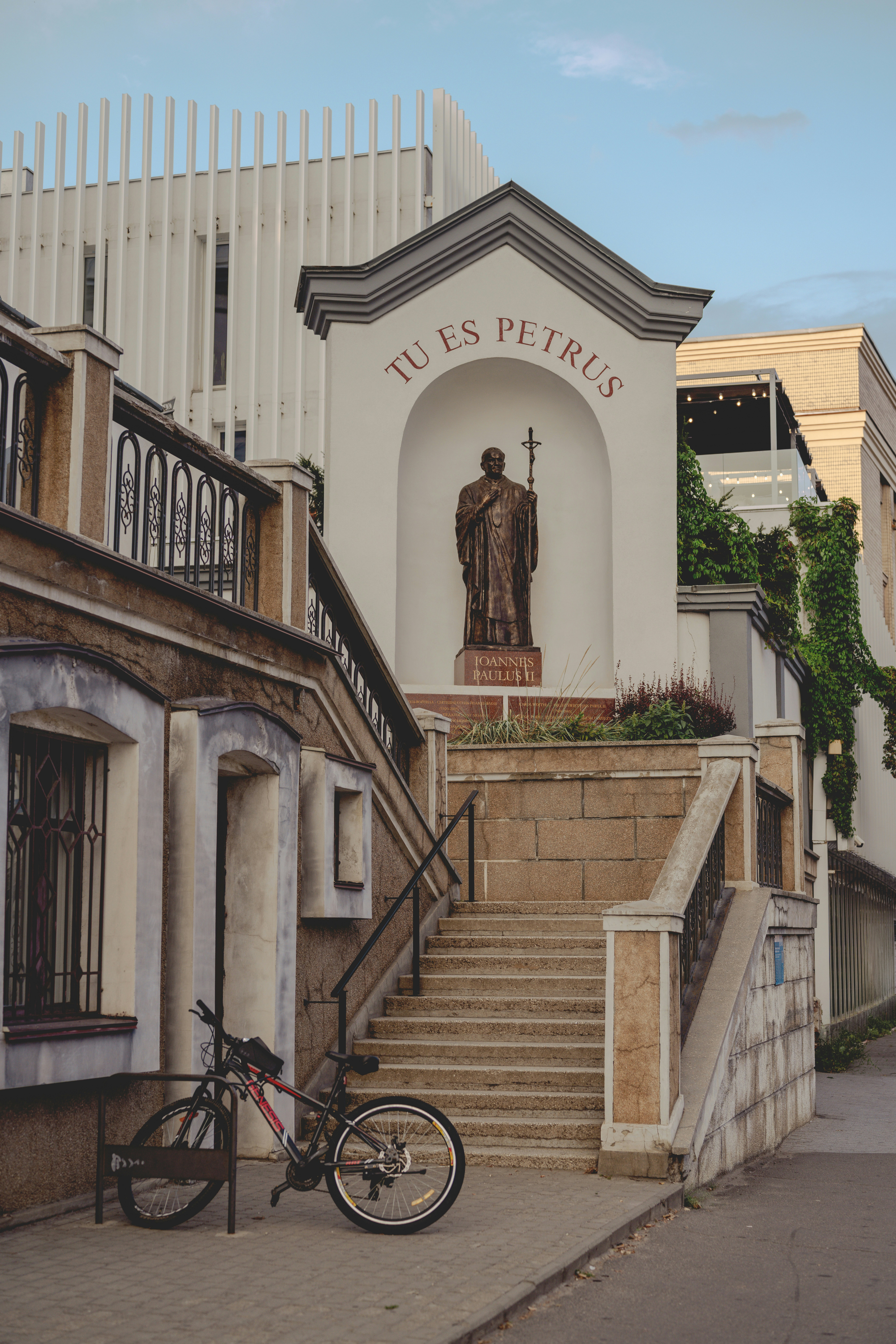 Statue of Saint Peter within an arched niche atop a stone stairway, flanked by a bicycle and urban architecture; a quiet street corner framed by warm exterior tones.