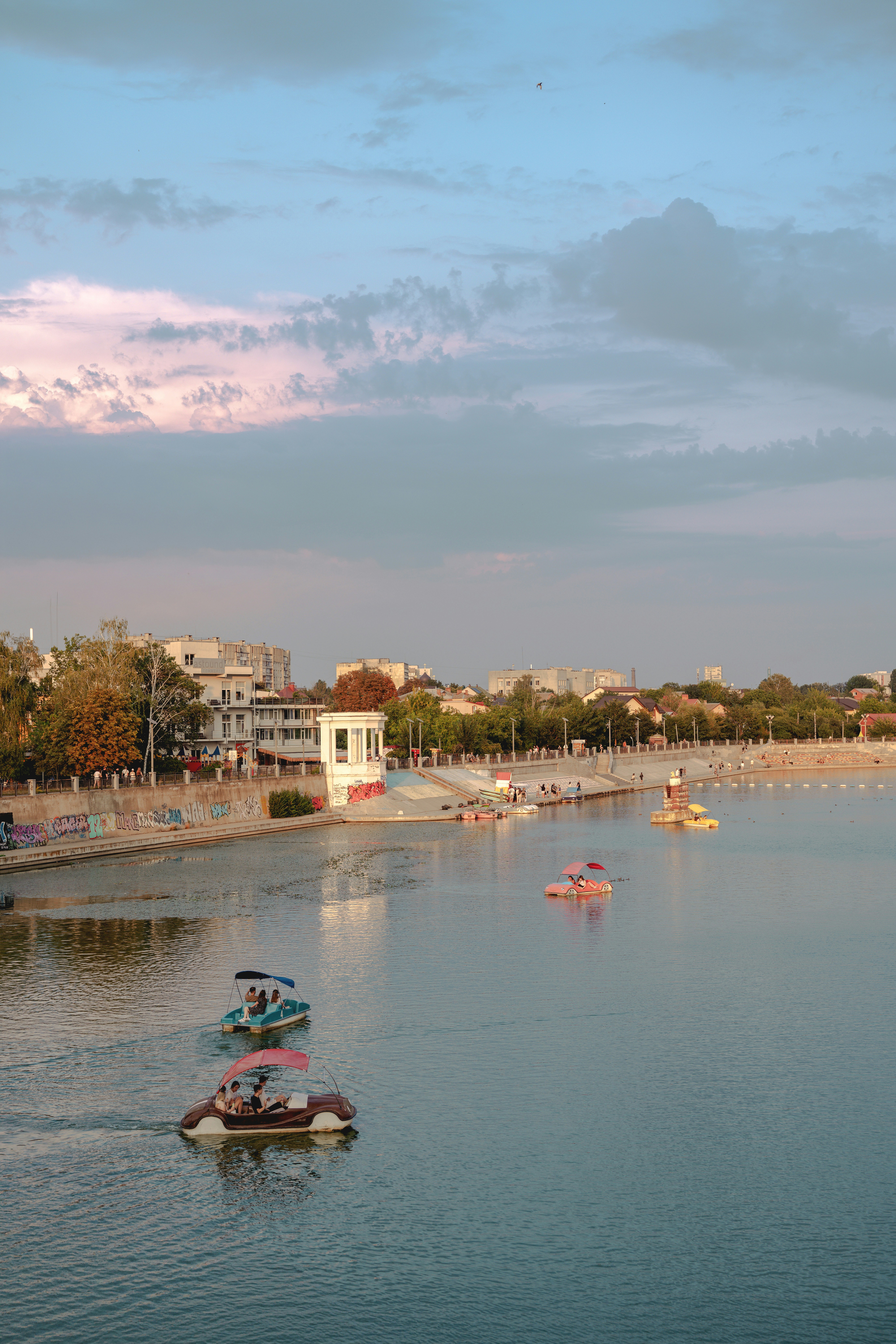 Sunlit riverfront with pedestrian promenade and pedal boats at dusk