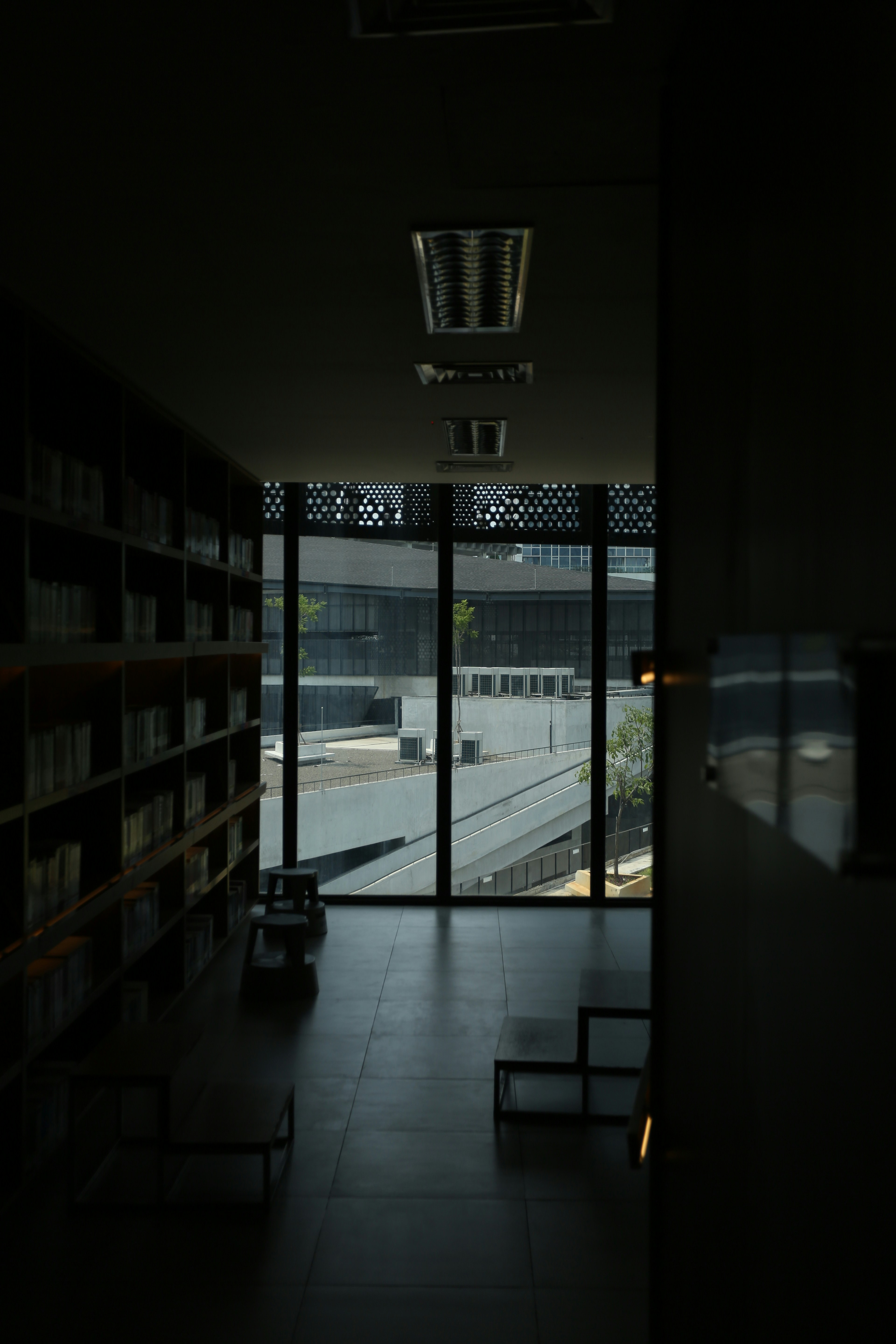 Interior of a dimly lit library or study hall with shelves on the left and large windows at the far end revealing an outdoor structure; the scene emphasizes depth and architectural lines through the corridor toward daylight.