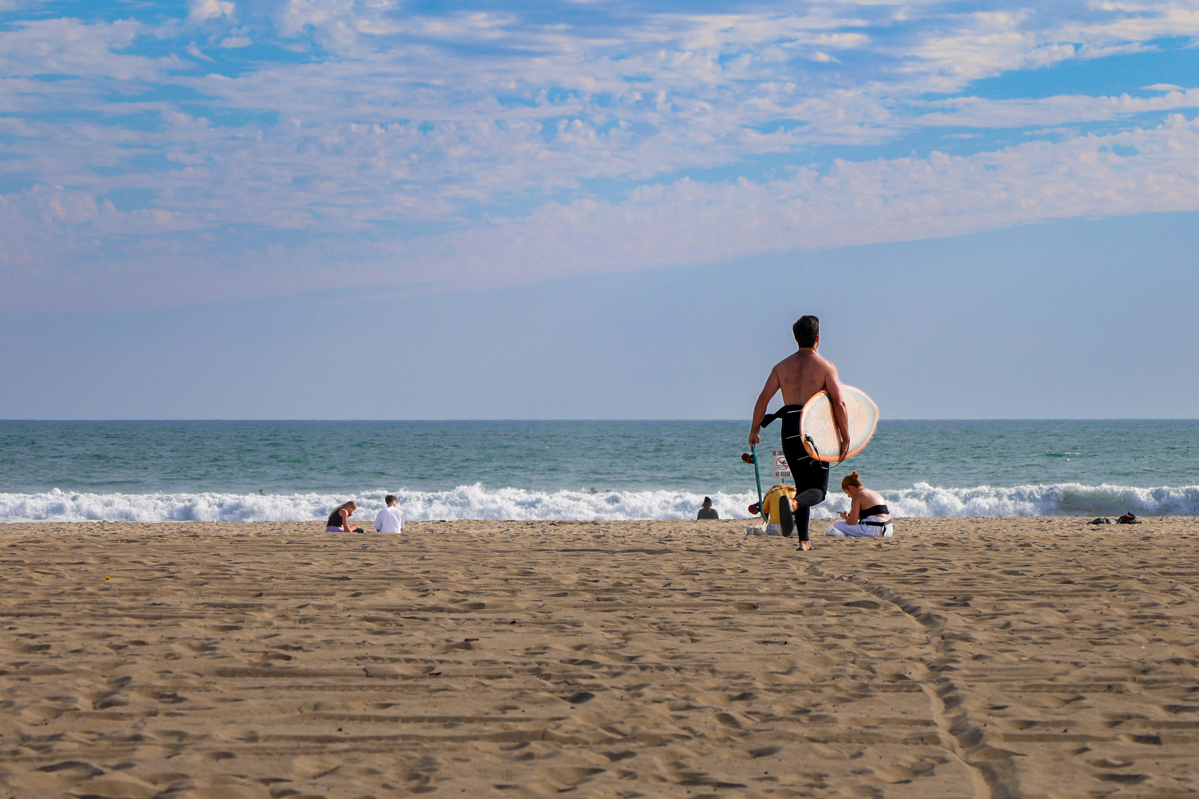 A lone surfer with a white board crosses a sandy beach toward the water, while a few beachgoers relax near the surf. The scene captures a calm moment at the coastline.