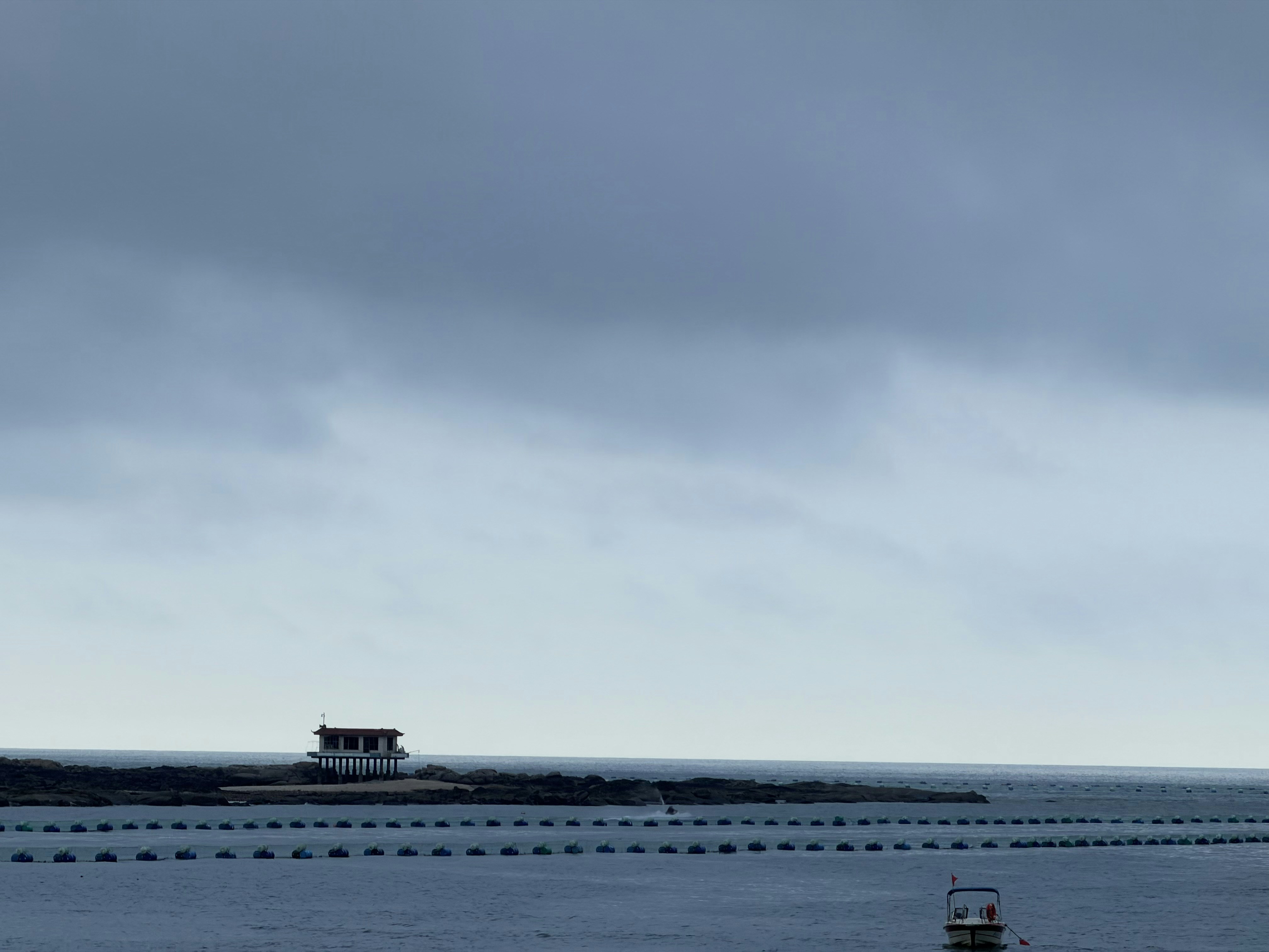 Distant shoreline with a small structure on the breakwater under a moody sky; a calm sea and repeating buoys create depth.