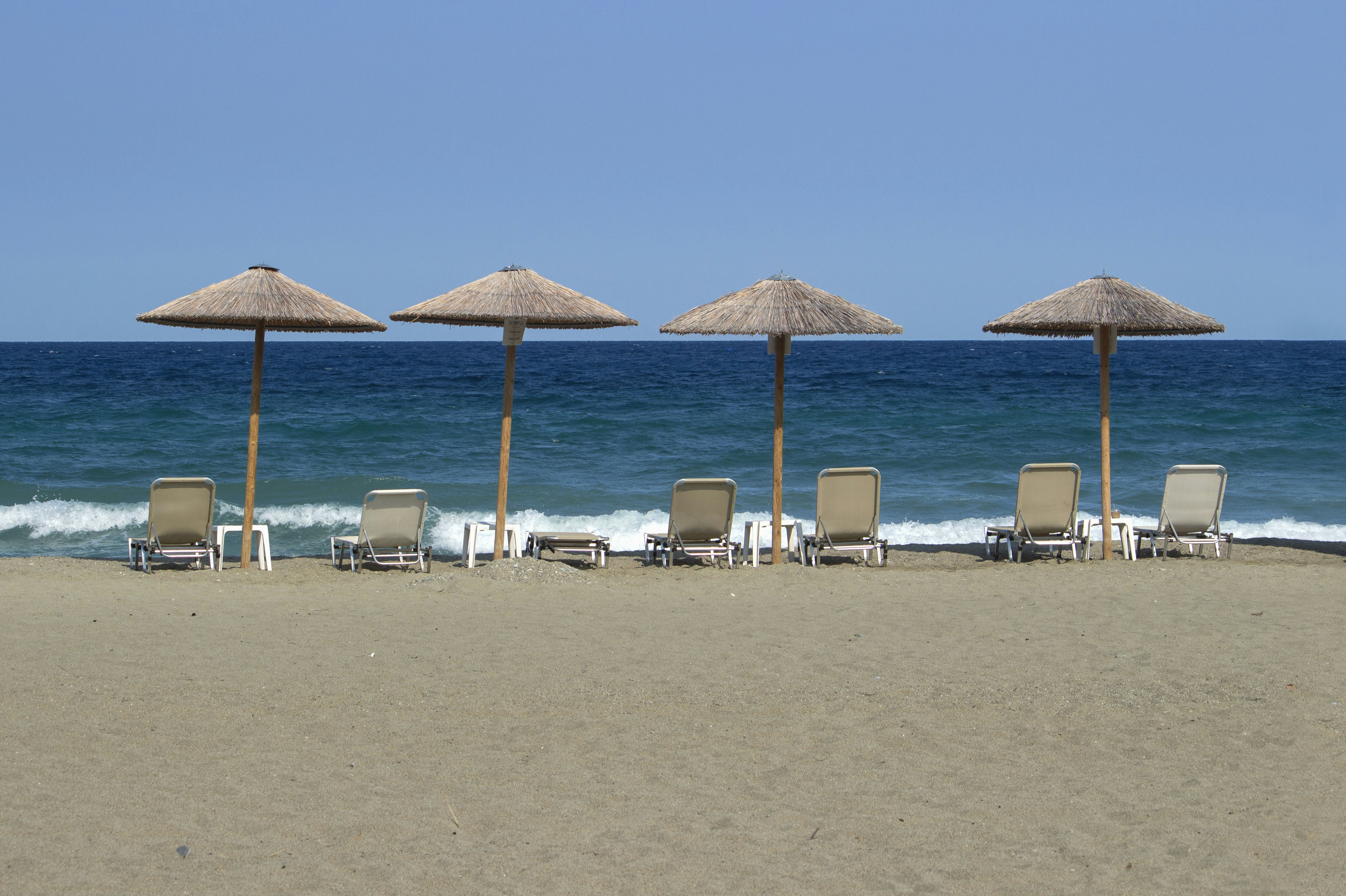 Rows of lounge chairs and thatched umbrellas along a sandy shore with calm blue sea beyond.