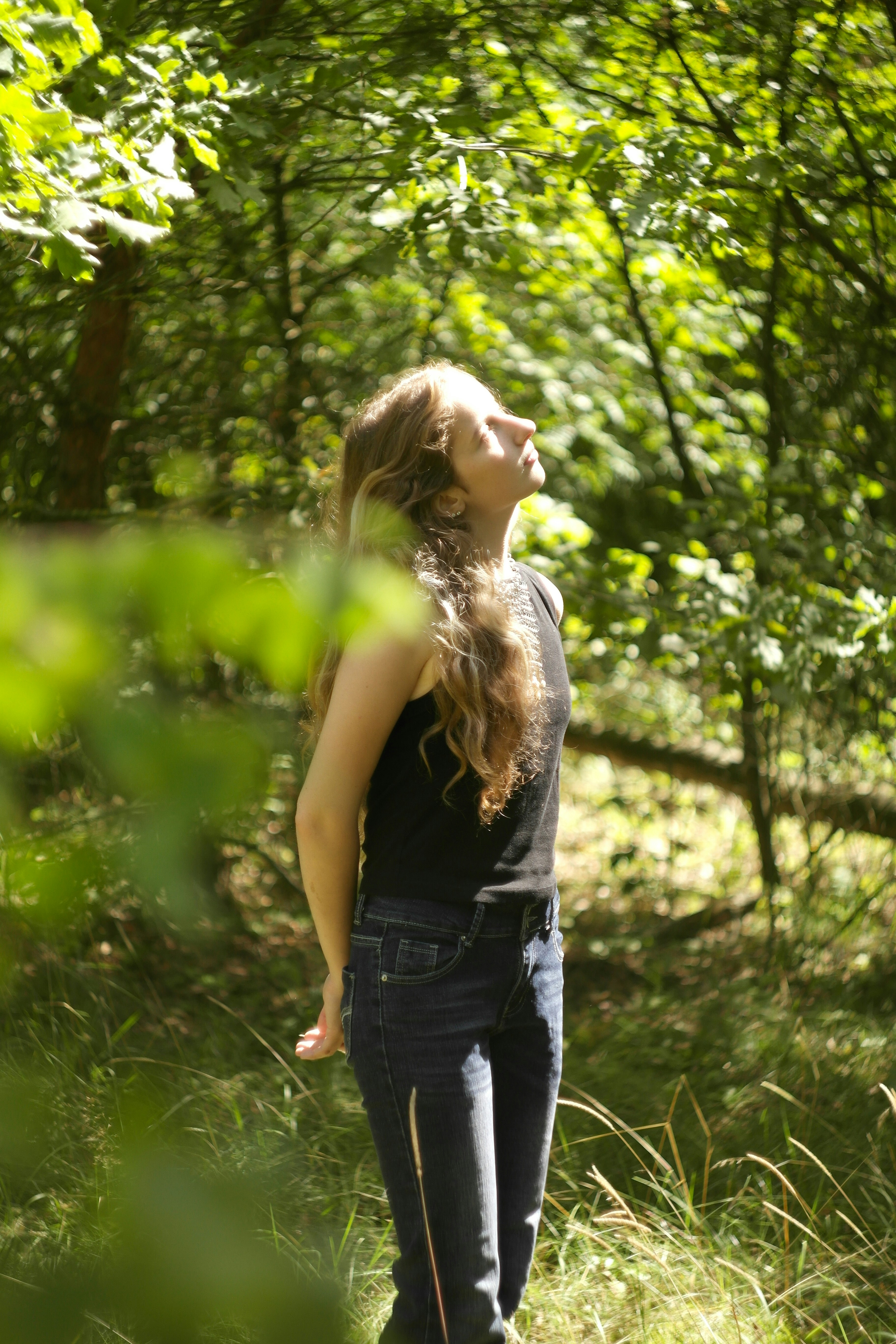 Sunlit girl stands in a sun-dappled forest clearing, wearing a black tank top and jeans, eyes closed as light pours over her face. A logo-free, candid outdoor portrait with soft bokeh from surrounding foliage.