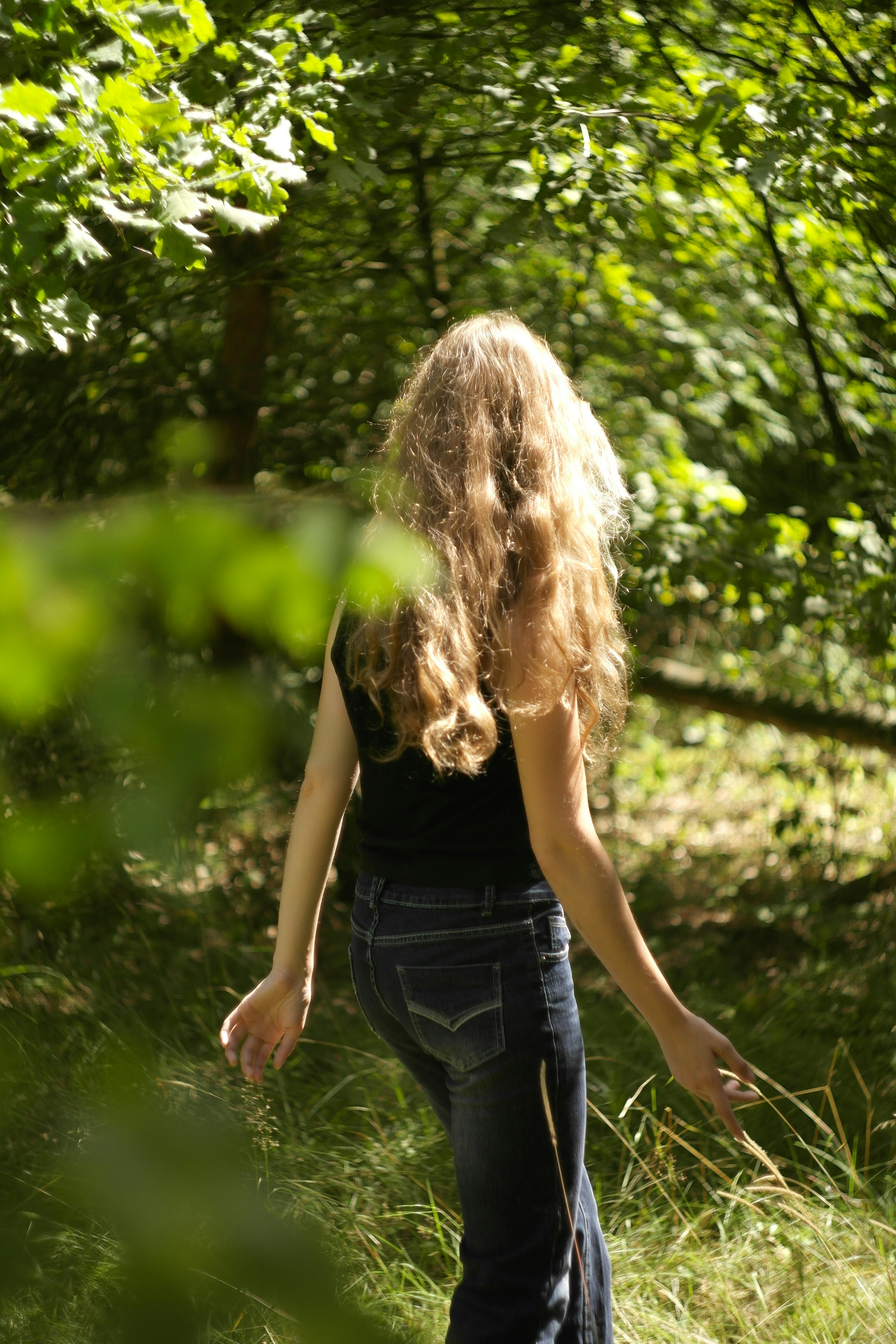 Sunlit back-portrait of a girl with long, wavy hair in a forest, wearing a black sleeveless top and jeans; the scene emphasizes natural light filtering through leaves and a blurred foreground frame.
