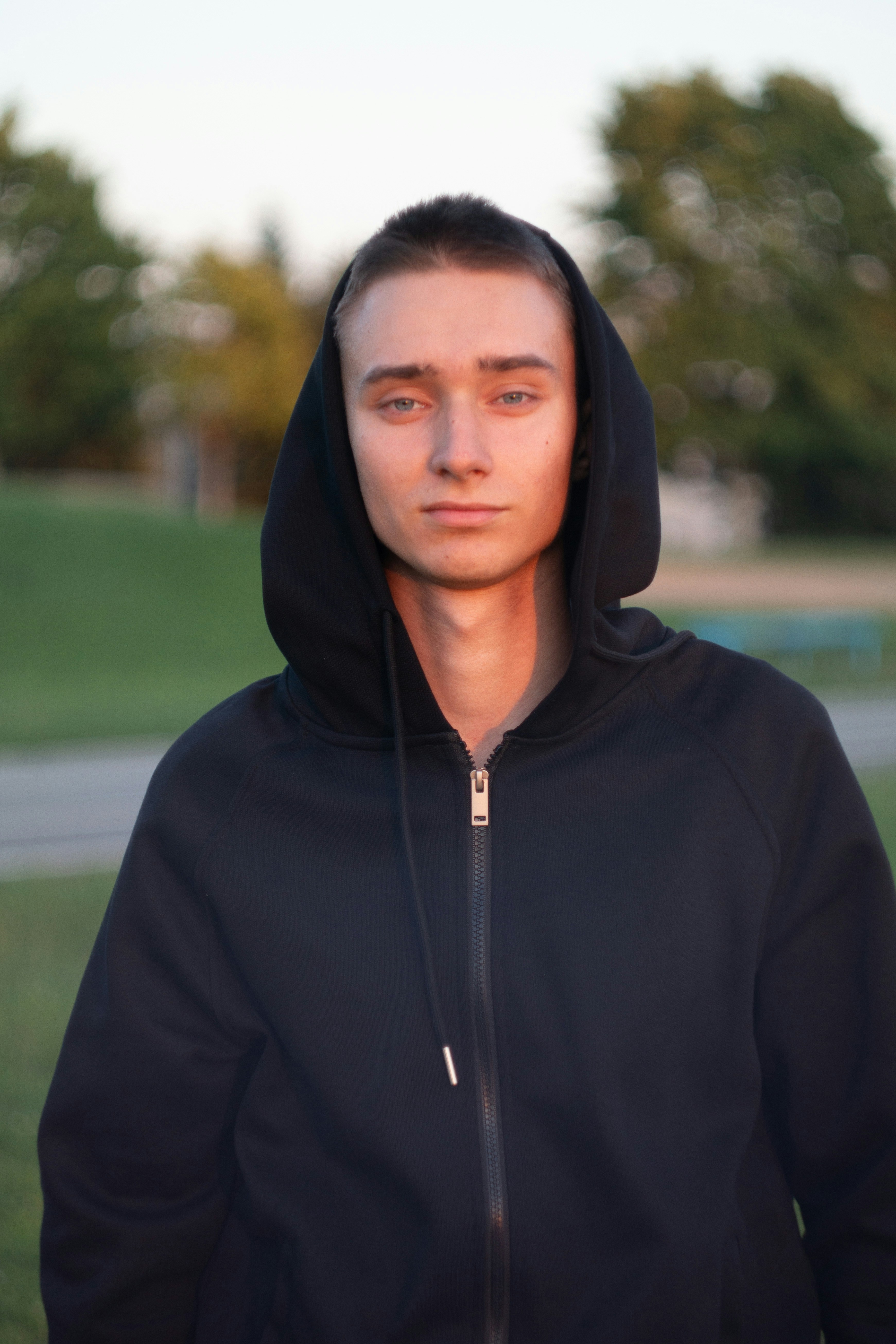 Young man in a black hoodie stares softly at the camera in a sunlit outdoor setting with blurred greenery in the background.