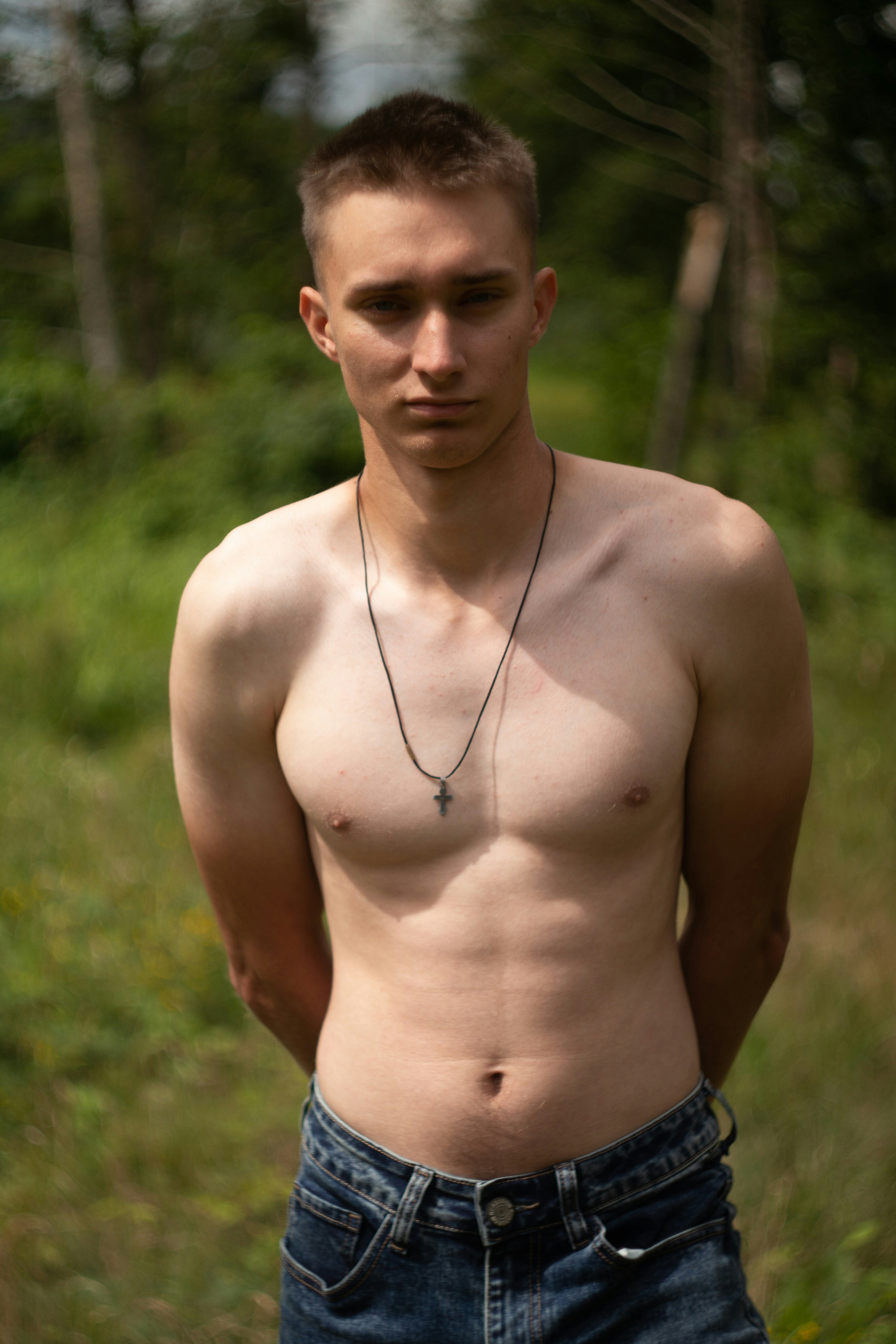 Shirtless young man stands in a sunlit forest clearing, arms behind back, wearing a simple necklace and jeans. A natural outdoor portrait emphasizing torso and facial expression.