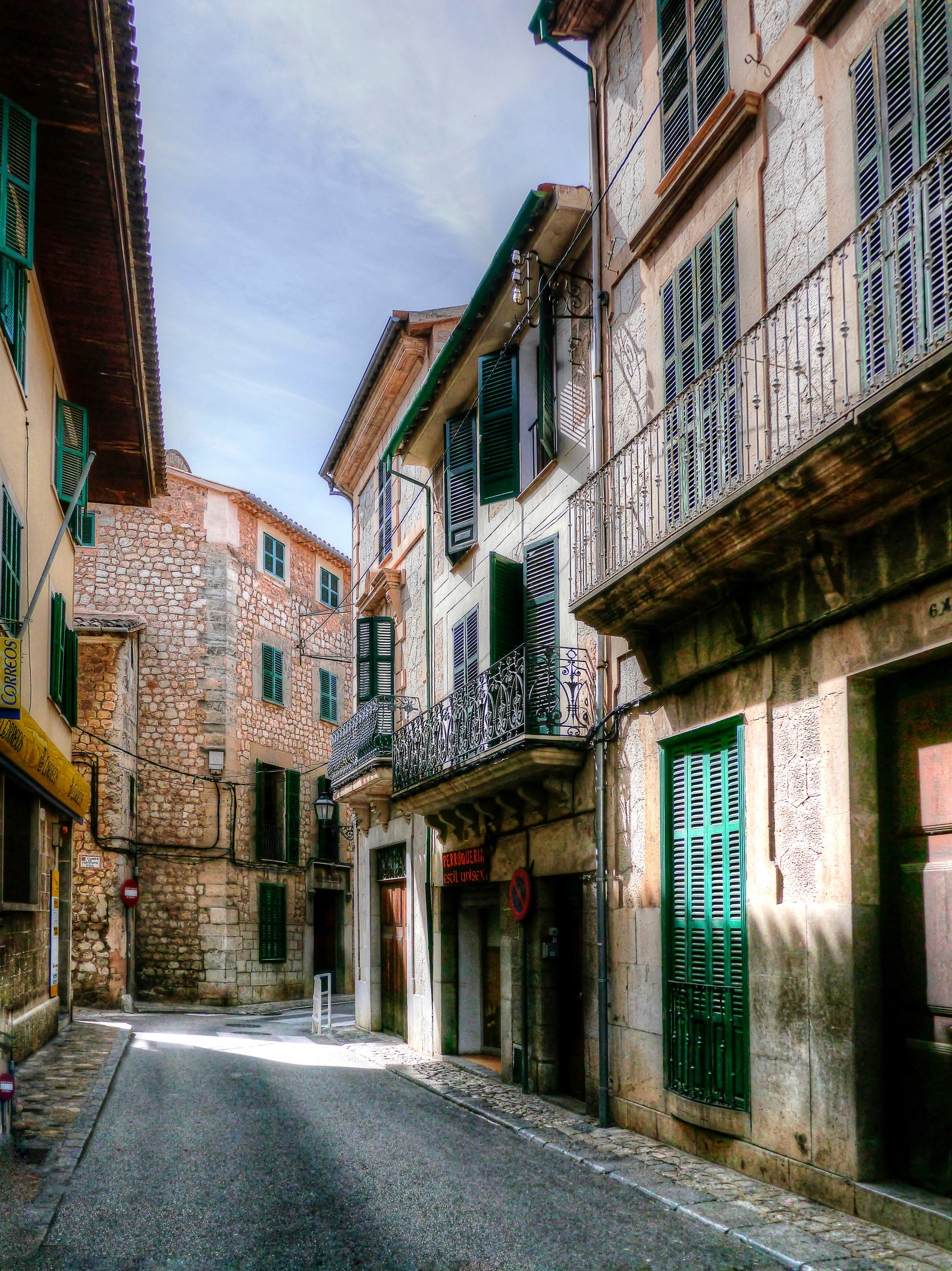 Stone-and-plaster street bends through sunlit history, flanked by balconies and green shutters on old town buildings.