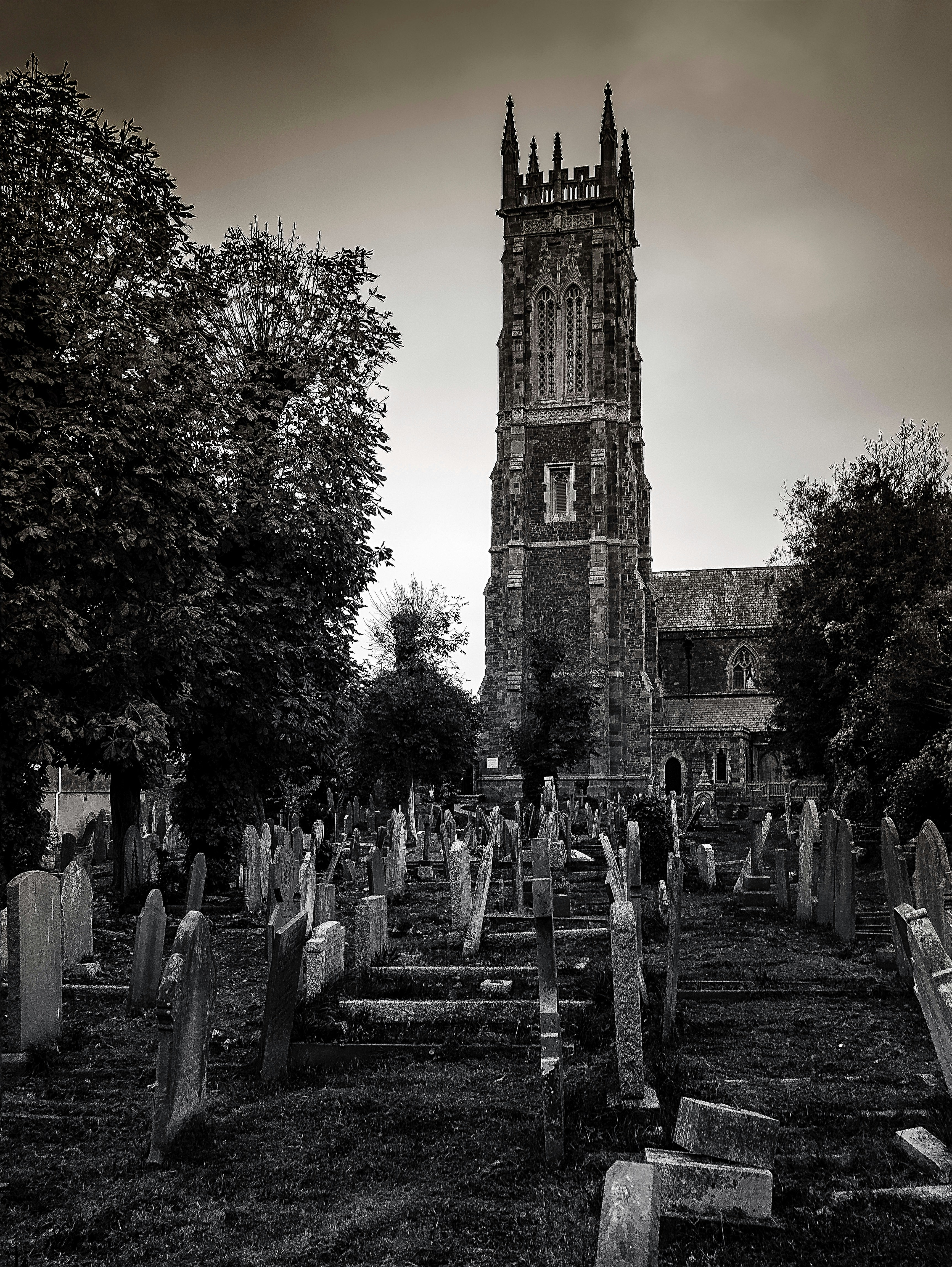 Monochrome cemetery with a tall church tower looming over weathered gravestones, captured with moody lighting and with the scene composed to emphasize vertical architecture and age-old serenity.