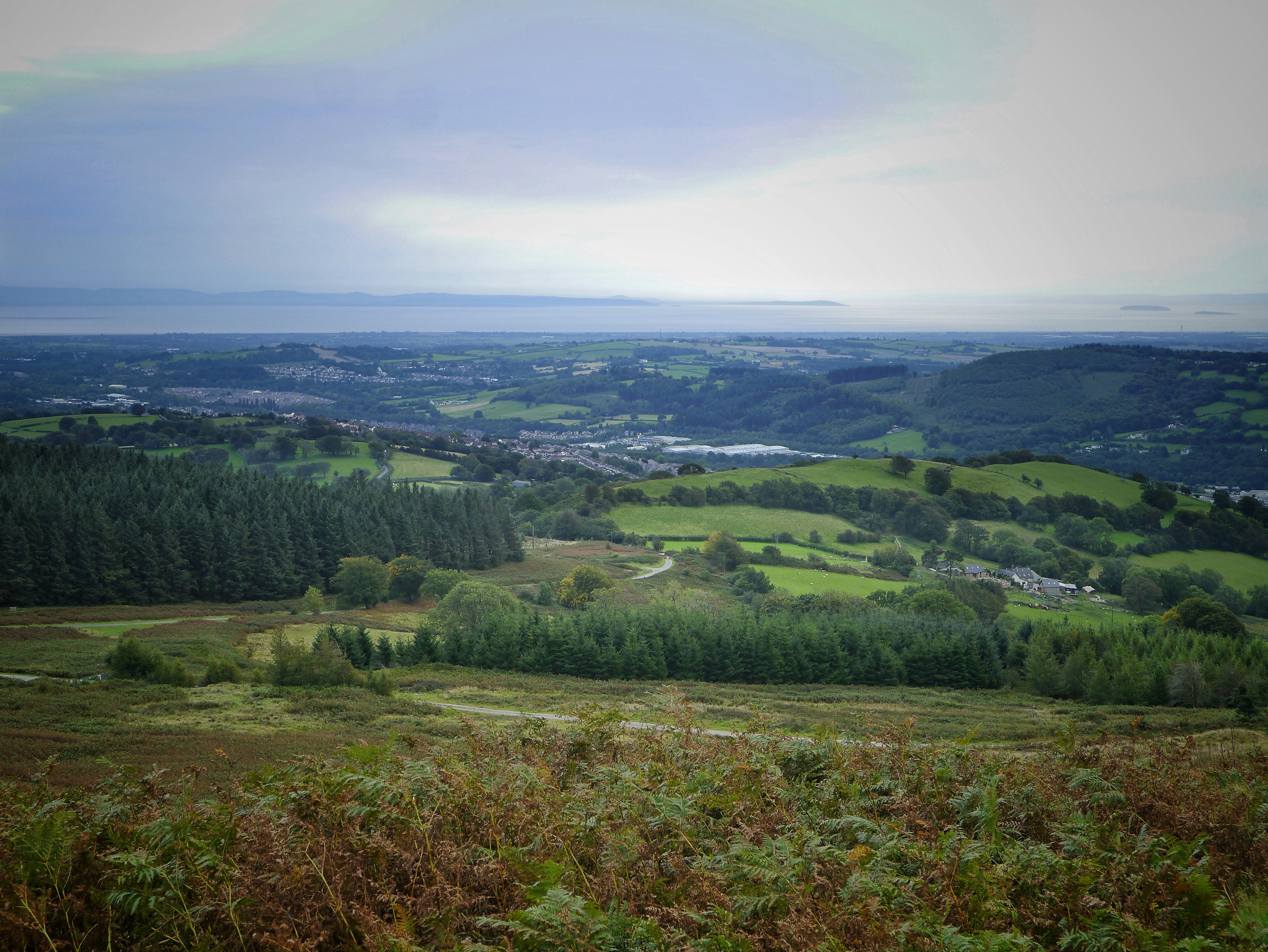 Rolling countryside valley with patchwork fields and distant hills under soft, overcast light; a tranquil landscape emphasizing layering of terrain and distant towns.