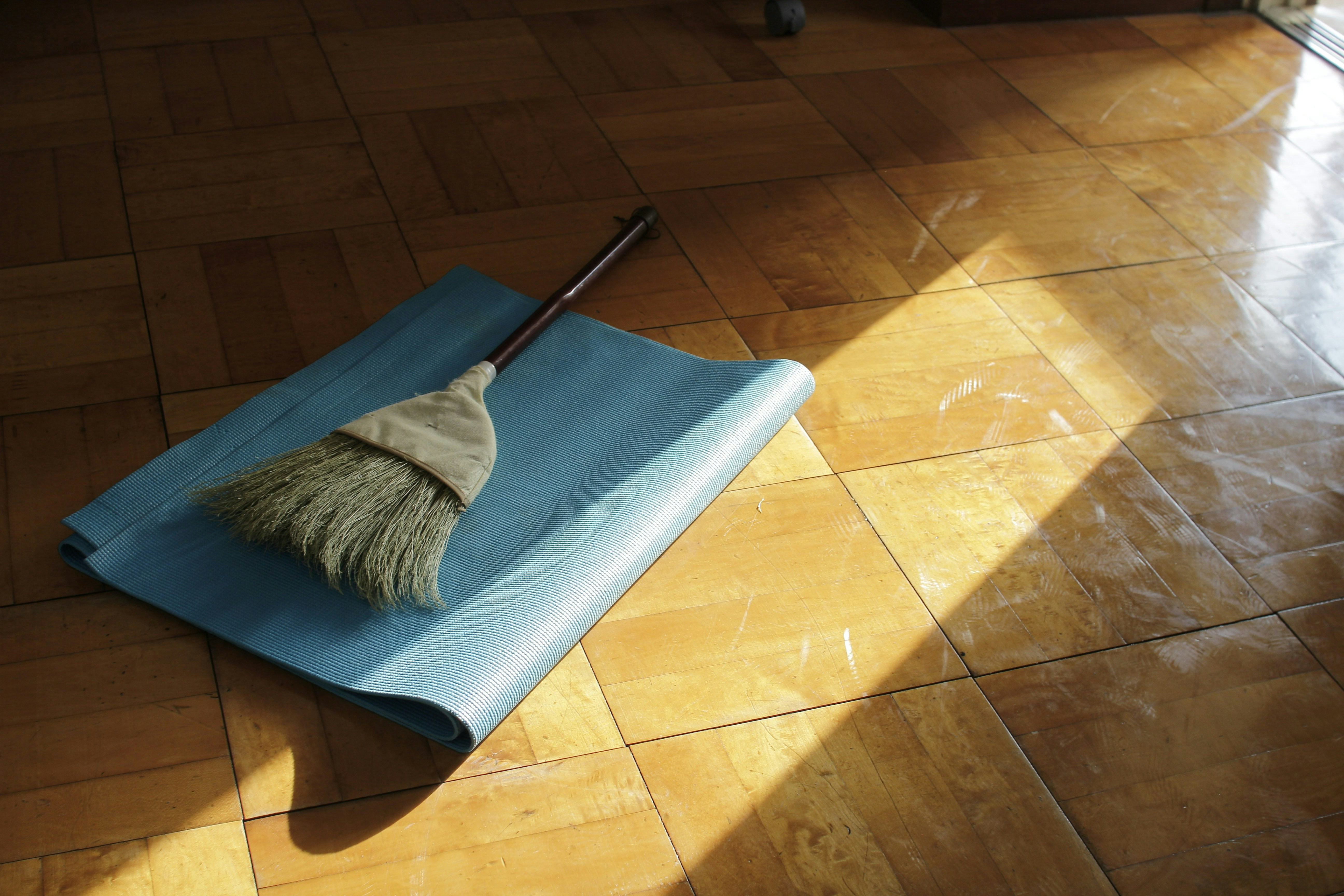 Blue mat with a painter's brush rests on sunlit wooden floor, a moment of simple domestic calm. The scene captures soft shadows and warm tones, emphasizing texture and everyday objects.