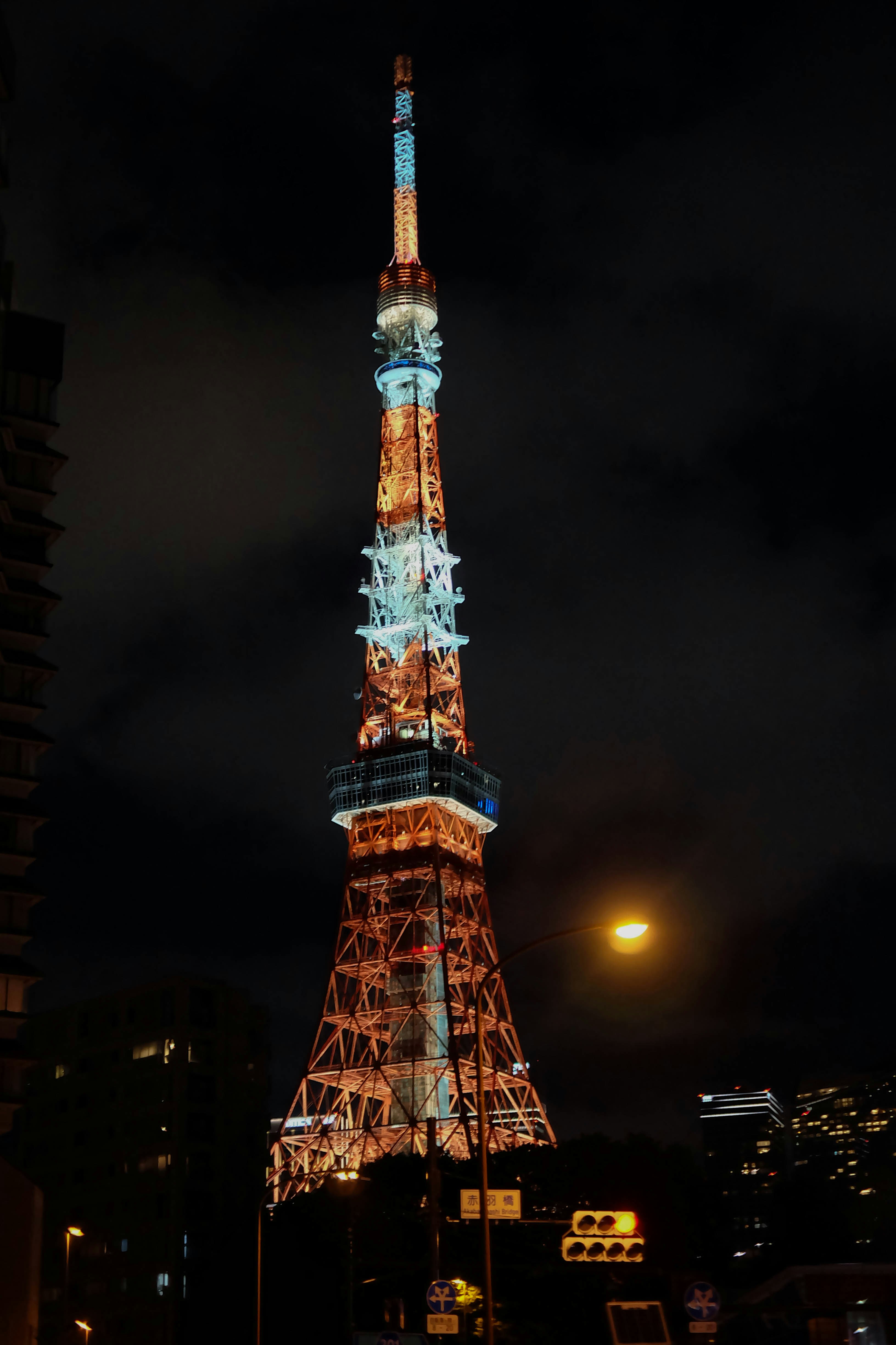 Night view of Tokyo Tower lit in warm orange and cool blue hues, rising over city lights and street lamps.