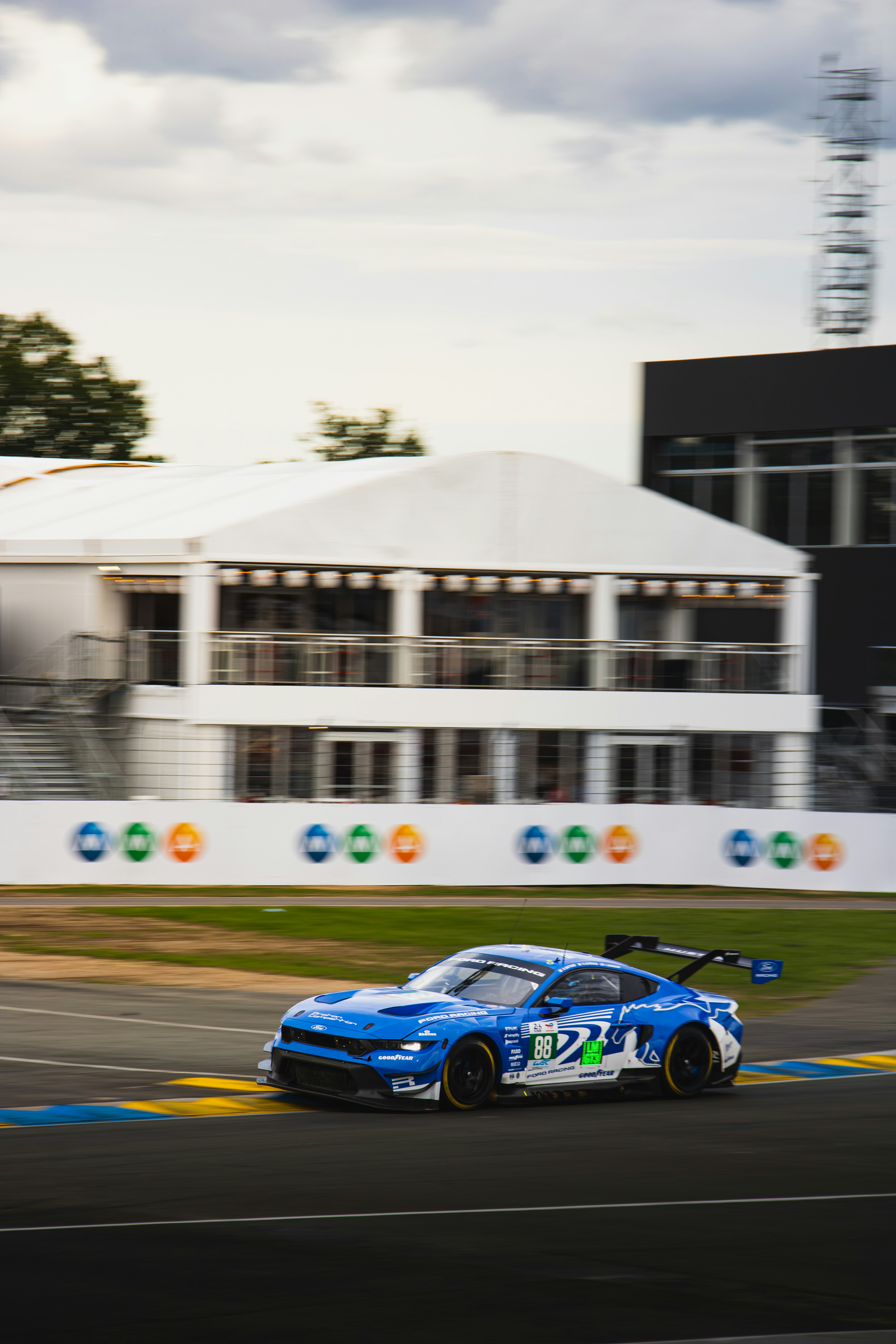 Blue race car speeds along track with blurred background and pit/stands in the distance; motorsport scene captured with motion blur. This is a photographic capture of a racing moment.