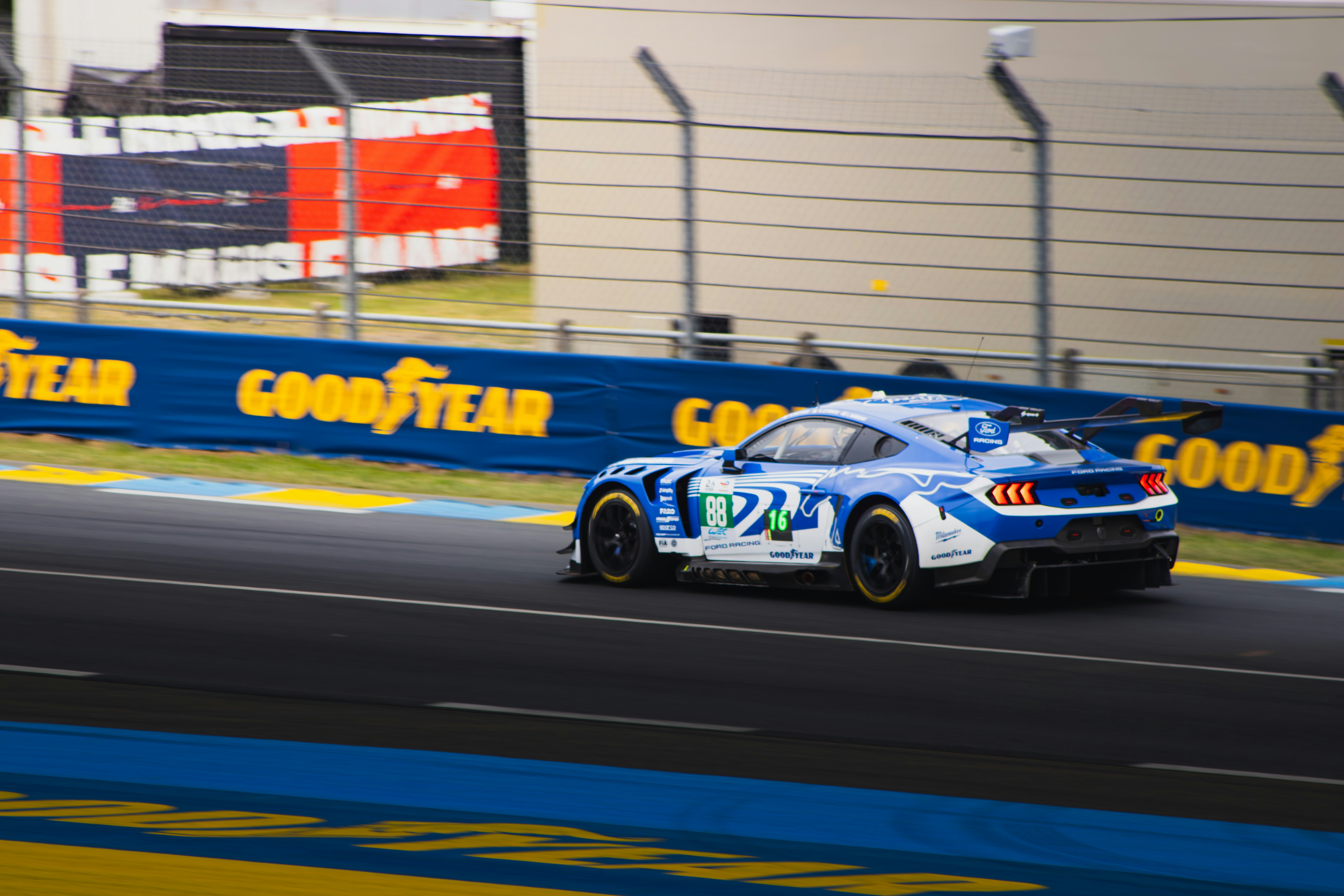 Blue and white race car with bold livery speeding along a track surrounded by Goodyear banners; motion blur emphasizes speed and competition.