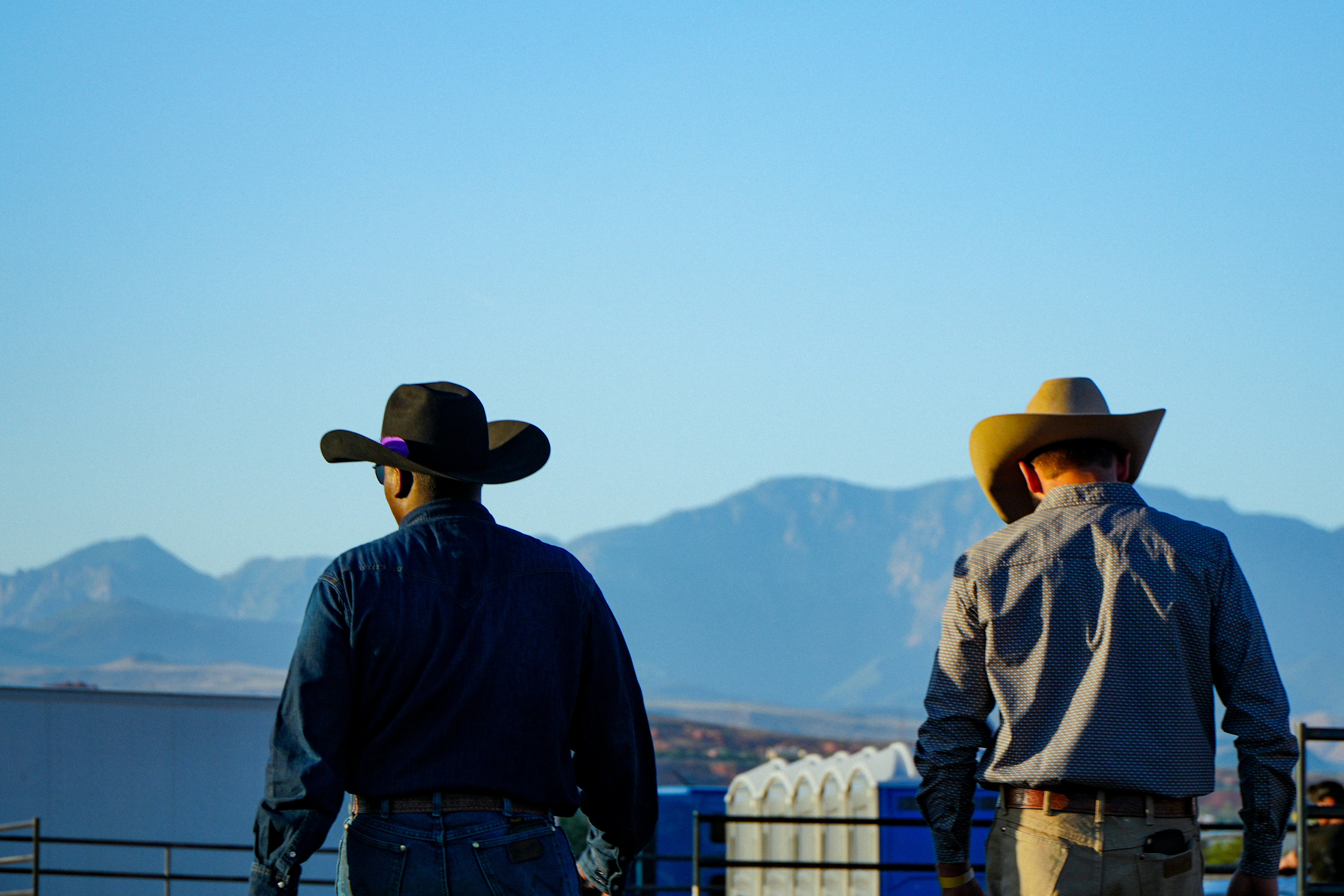 Two cowboys stand with backs to camera on a rooftop with mountains in the distance, under a clear blue sky.