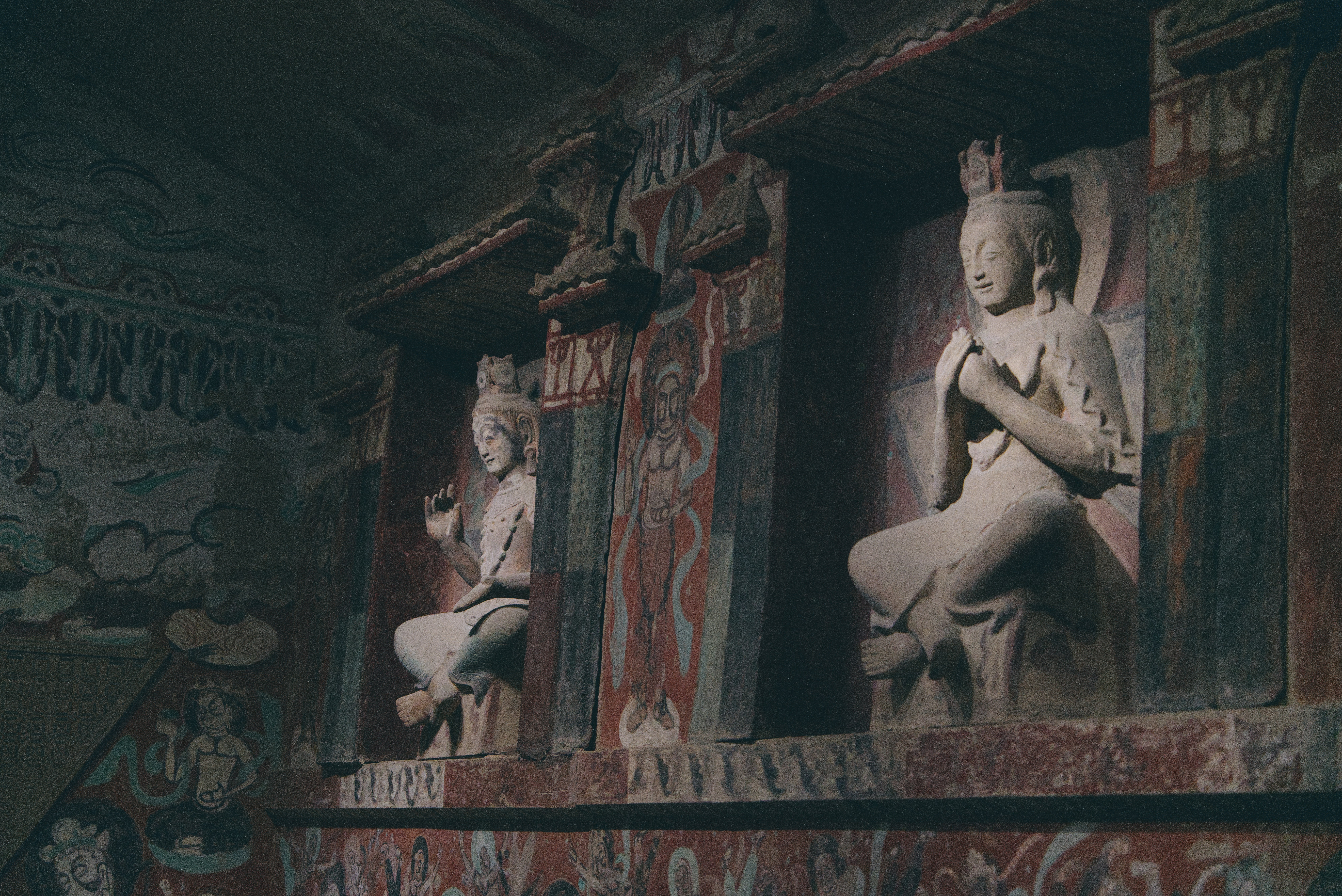 Dimly lit temple reliefs show seated stone Buddhas within a red painted corridor, carved figures framed by ornate painted columns and murals.