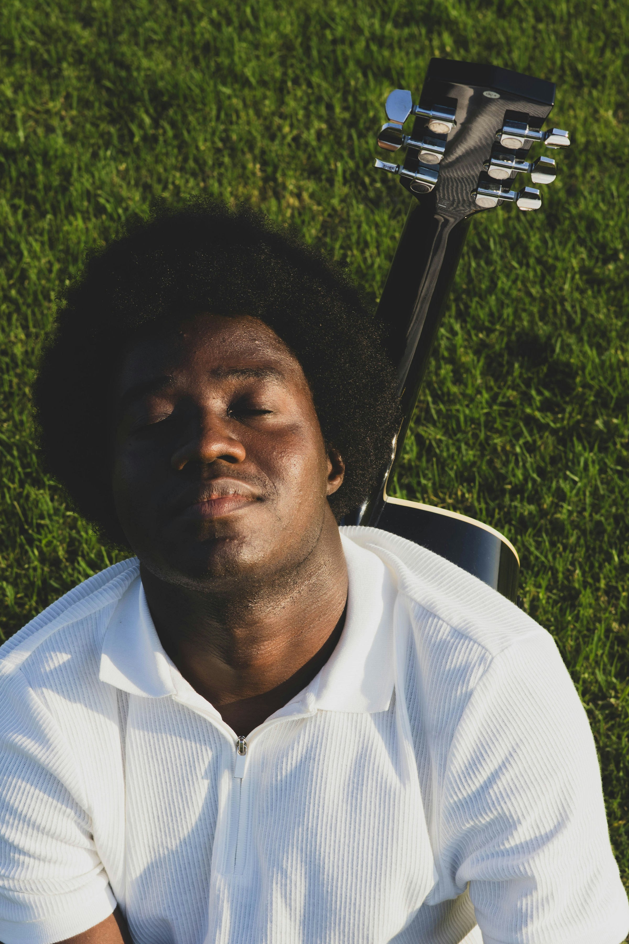 Portrait of a man lying on grass with a guitar resting beside him; a calm, sunlit moment highlighting relaxed pose and textured skin. (photograph)