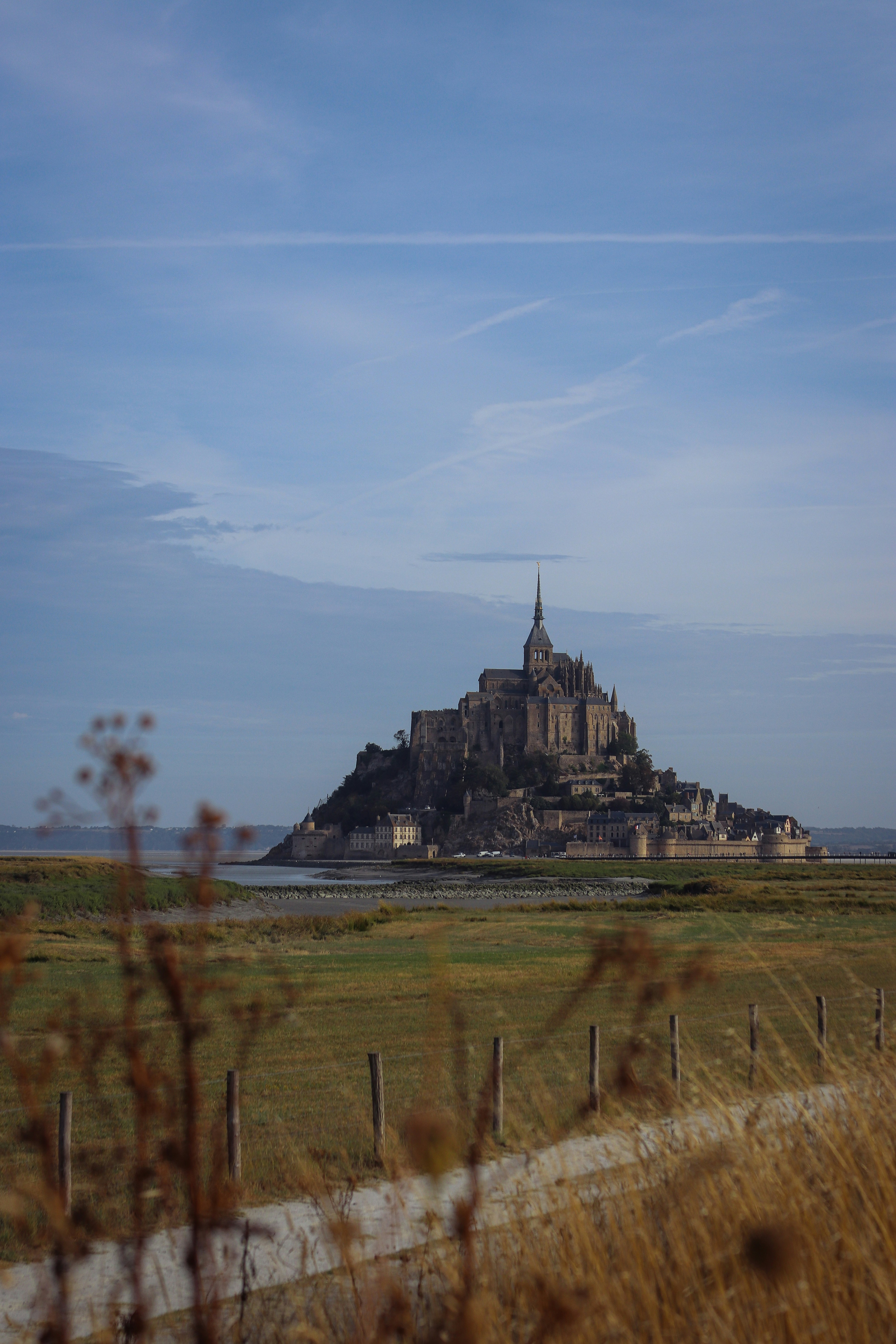 Towering medieval abbey on a rocky island framed by a low-horizon field and distant water under a blue sky.