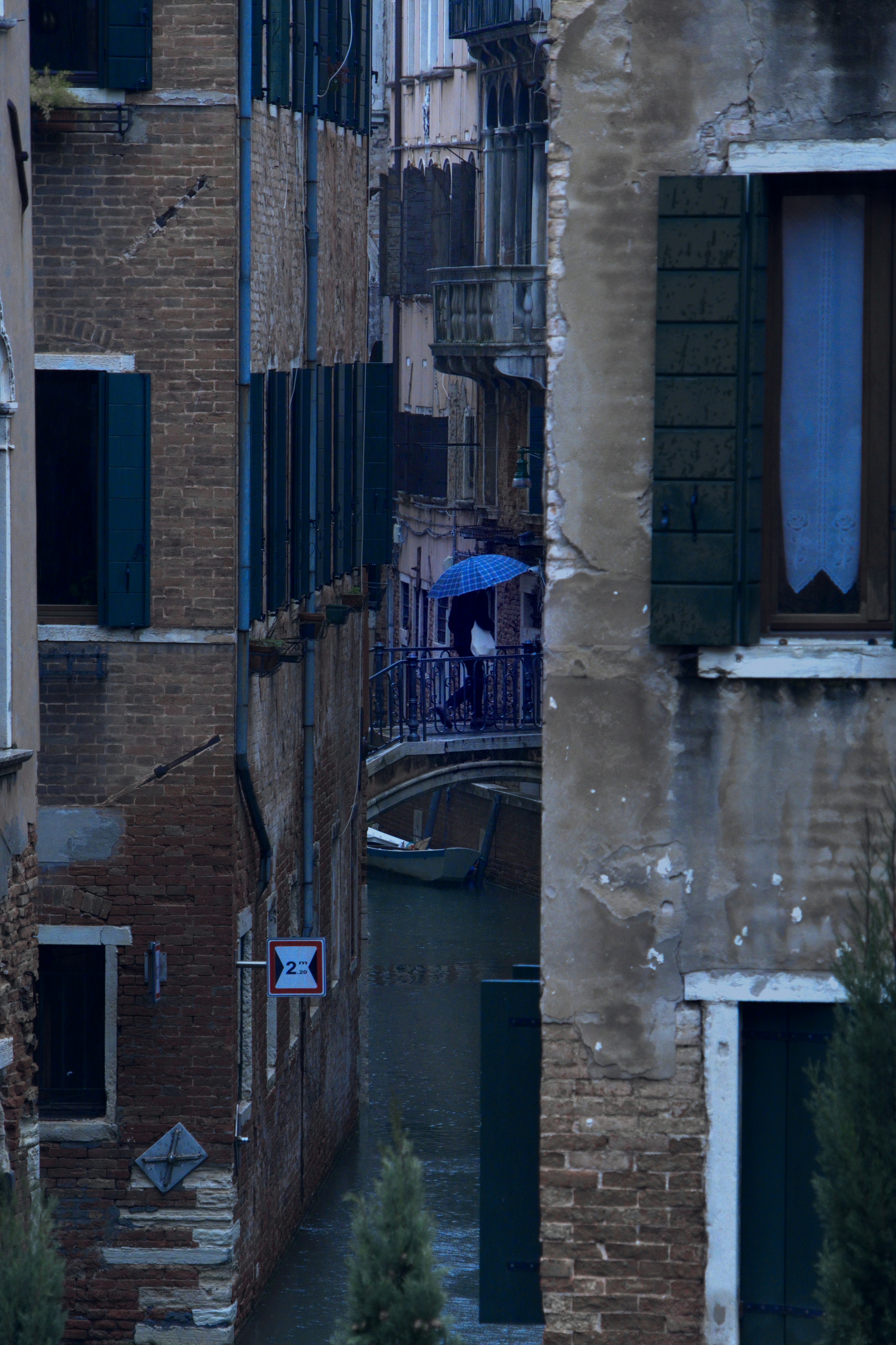 Narrow canal street with old brick buildings and a solitary figure with a blue umbrella on a small bridge over water.