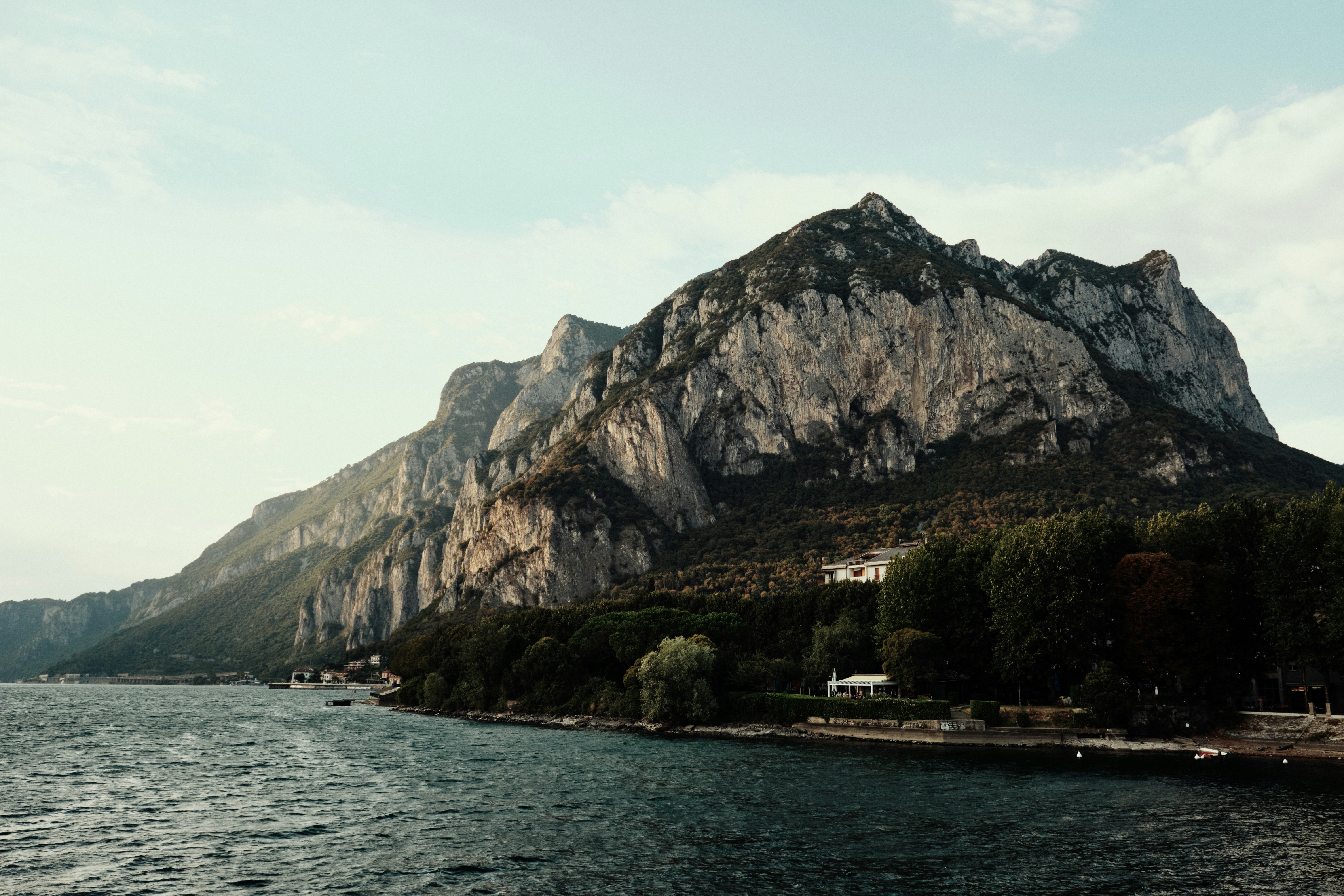 Coastal mountains loom over a quiet lake with a tree-lined shore and scattered buildings at the waterline. The scene emphasizes rugged rock, gentle water, and atmospheric light.