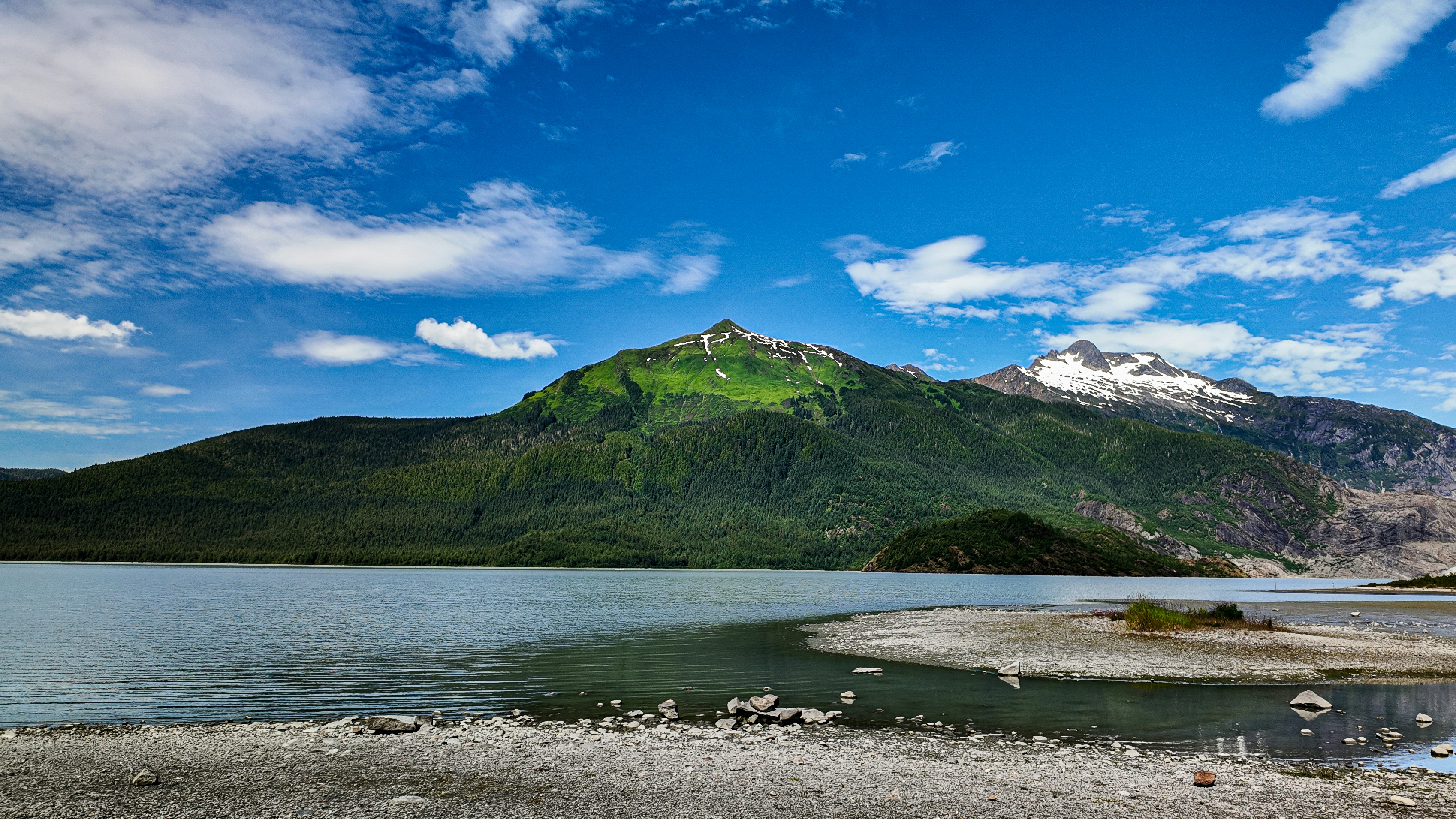 Green, pine-dotted mountains rise beside a calm lake with a rocky shore and a bright blue sky.