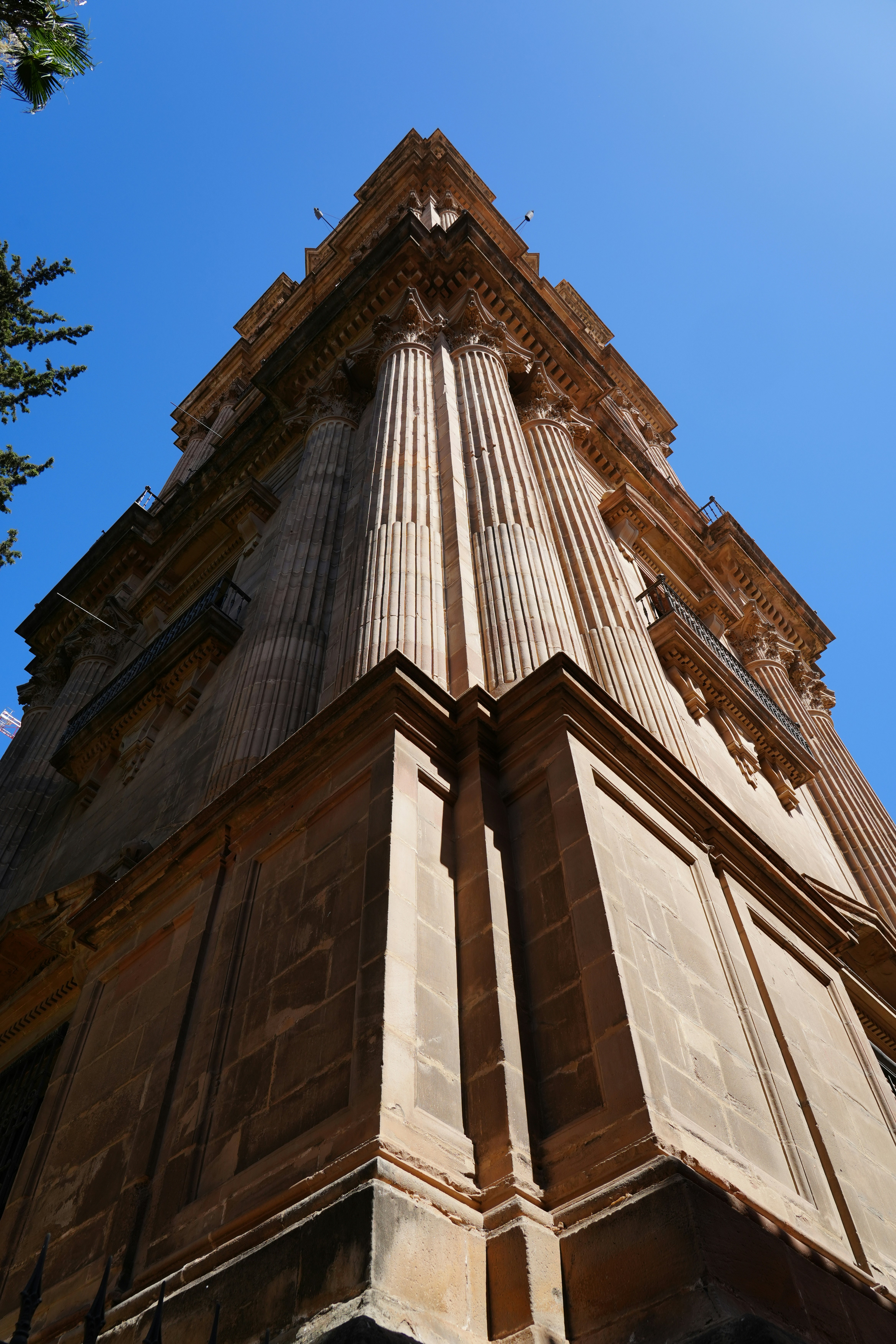 A towering ancient-style stone façade with fluted columns rises toward a clear blue sky, captured from a low angle to emphasize its vertical grandeur and architectural details.
