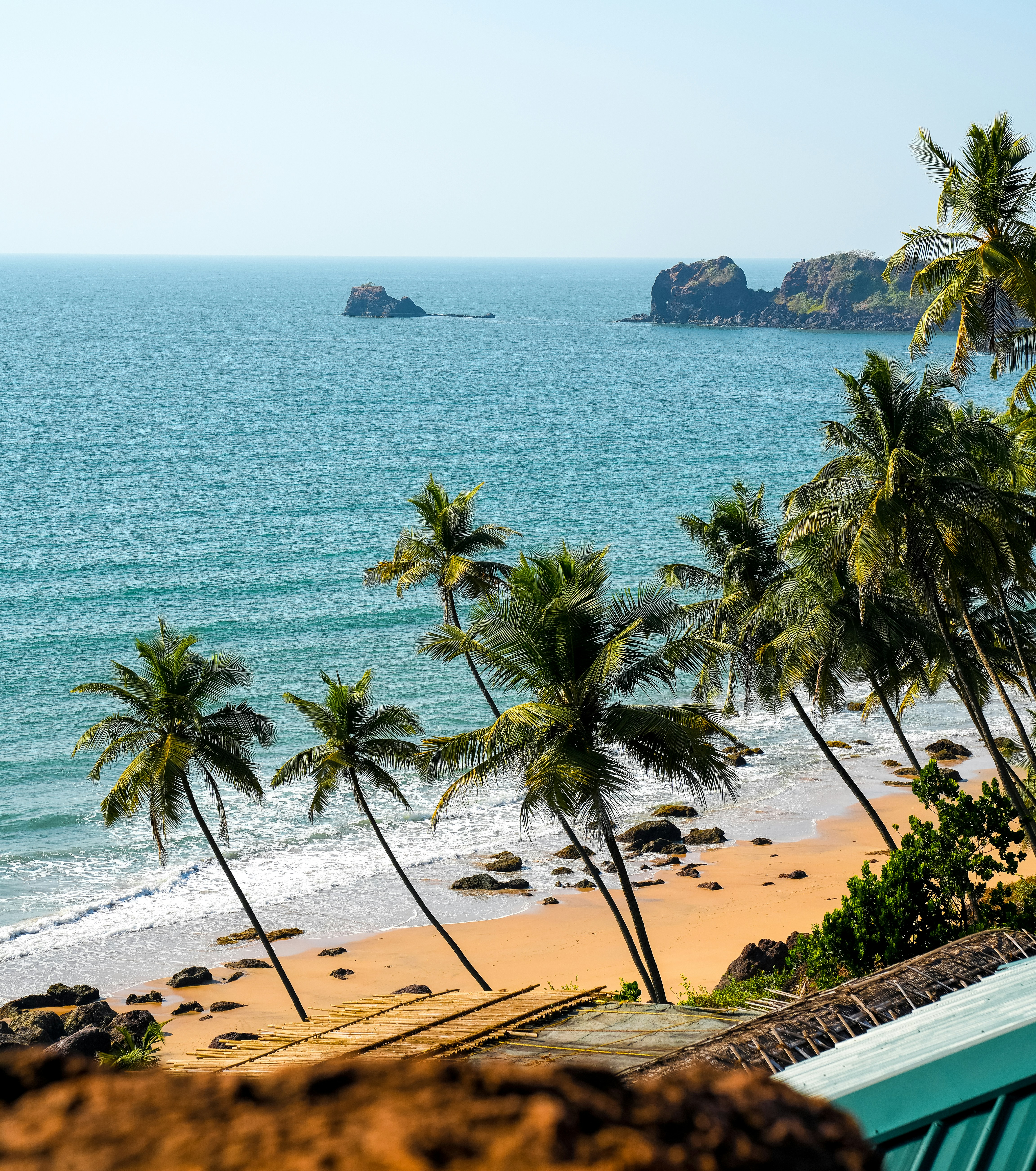 Sunny tropical beach with leaning palm trees along a sandy shore and turquoise sea, framed by distant rock outcrops and a turquoise railing in the foreground.