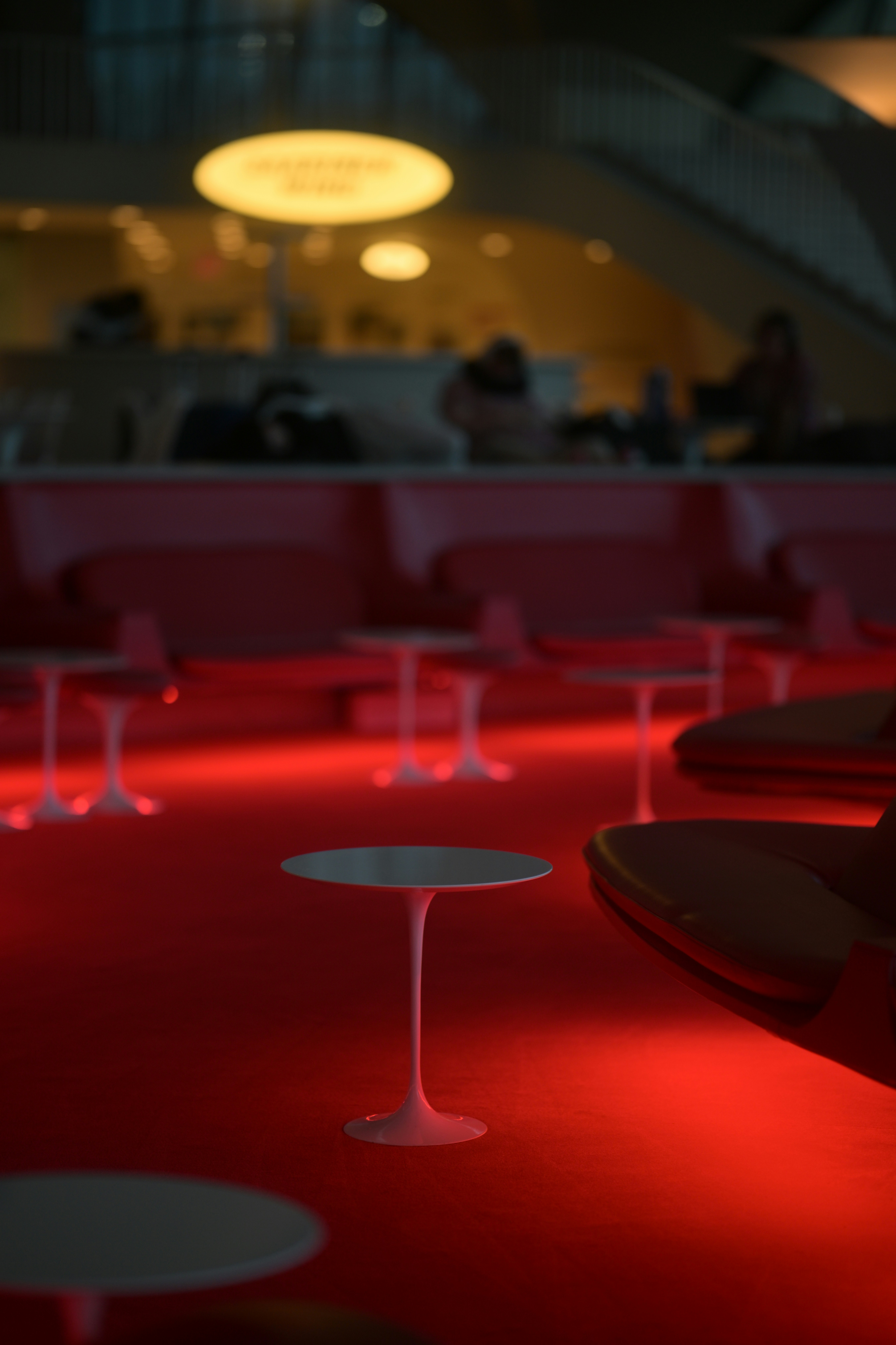A dim lounge scene with white pedestal tables bathed in red light, blurred seating in the background, and a glossy, curved surface reflecting the glow.