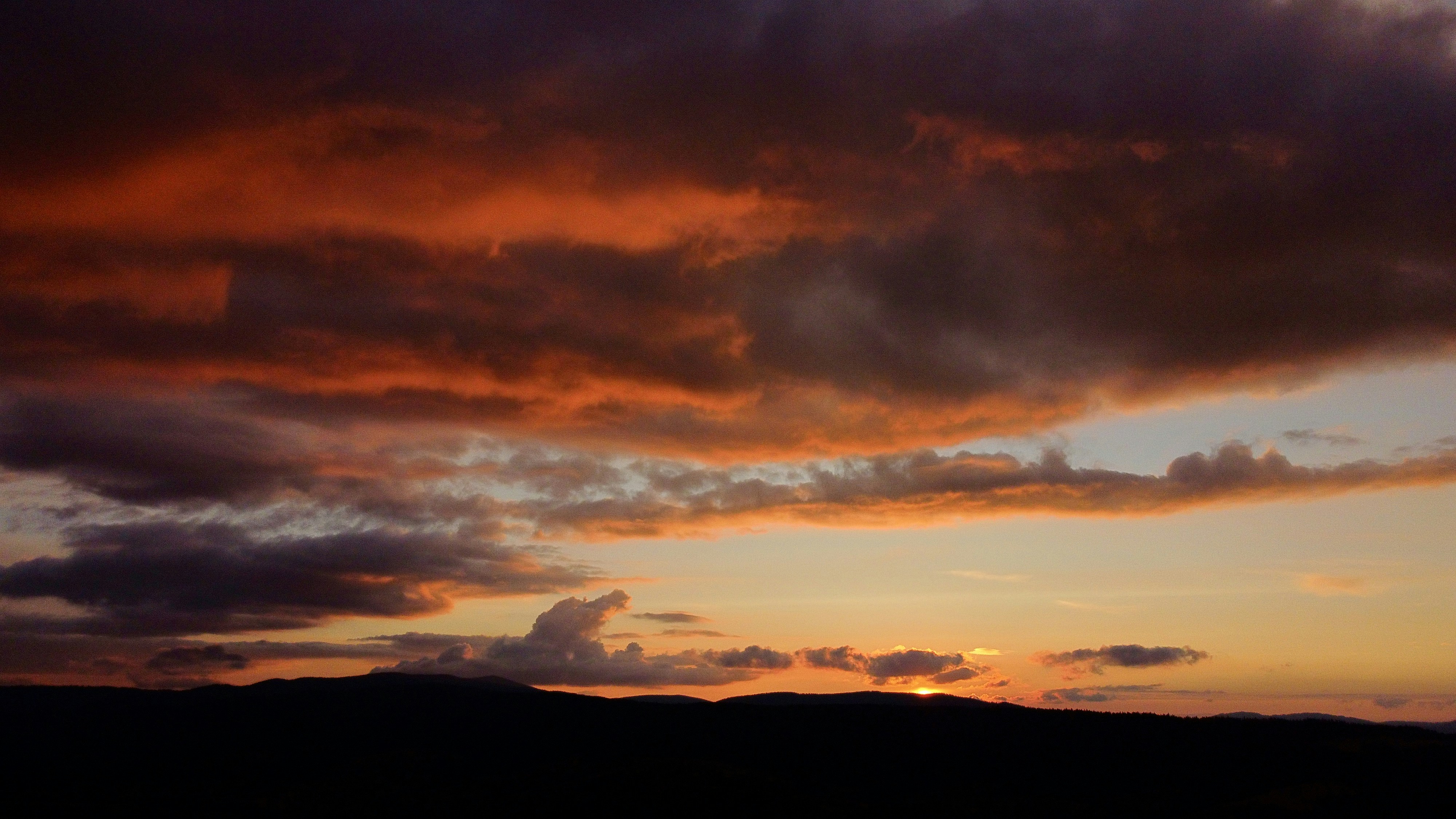 A low sun paints the horizon with warm orange and gold tones above a silhouetted ridge, clouds catching color as the landscape recedes into shadow.