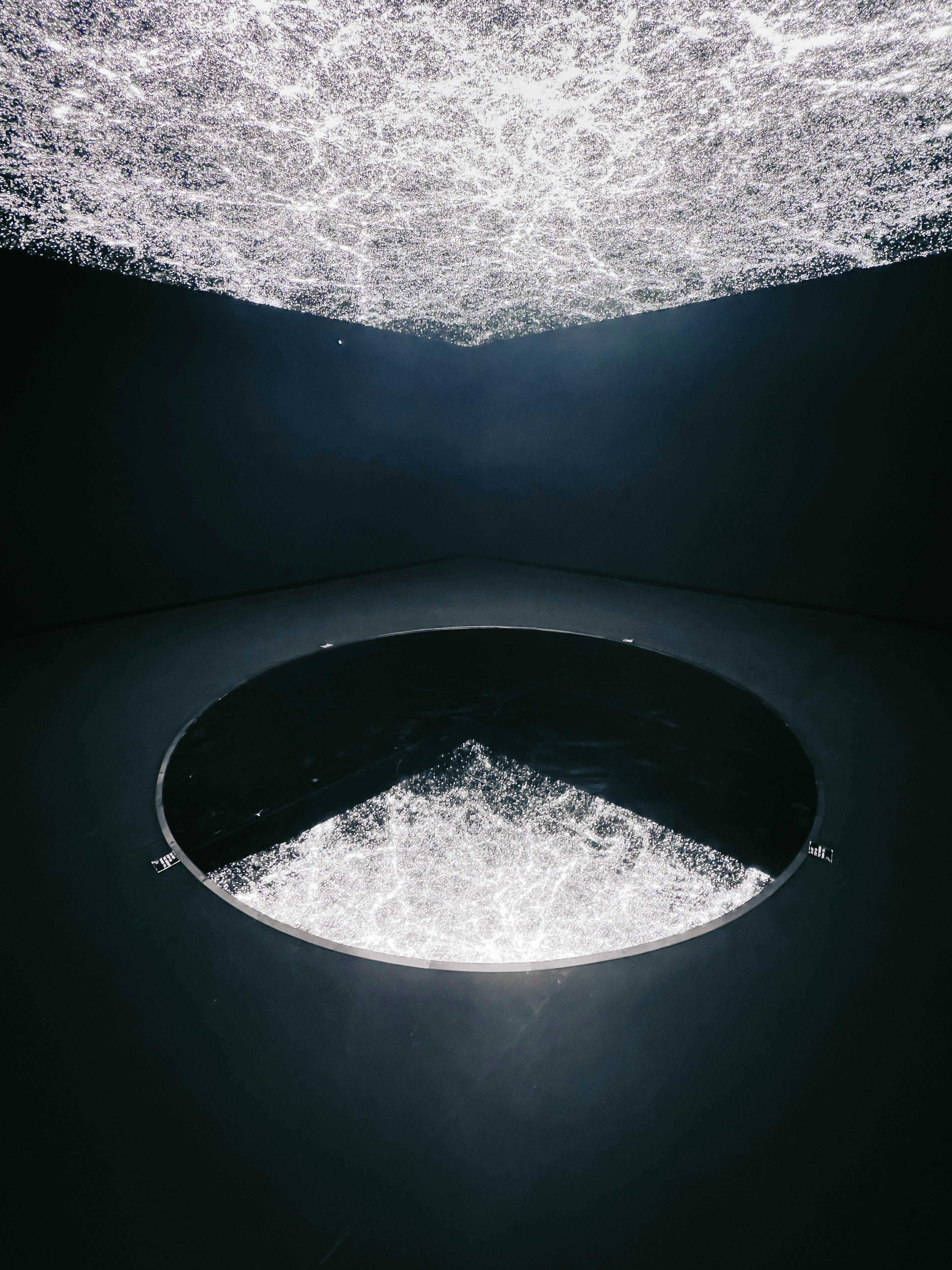Circular reflective disk in a dim room, with a bright, textured ceiling reflection forming a V-shaped focal symmetry. A minimal installation piece emphasizing light, shadow, and geometric balance.