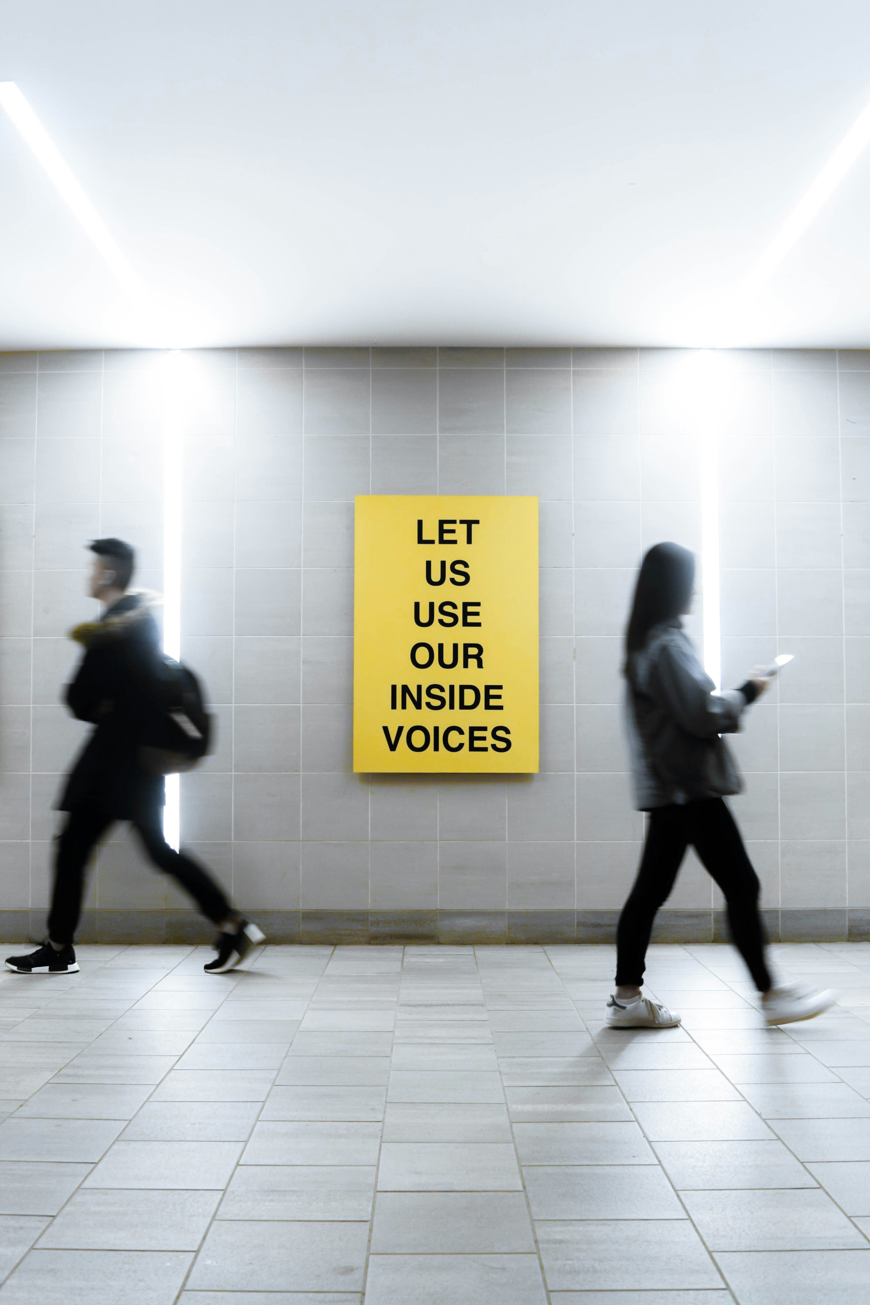 Yellow poster mounted on a tiled wall in a well-lit subway-like corridor, with two blurred figures walking past. The scene emphasizes motion around a central, legible message; the work is a graphic print in a public space.