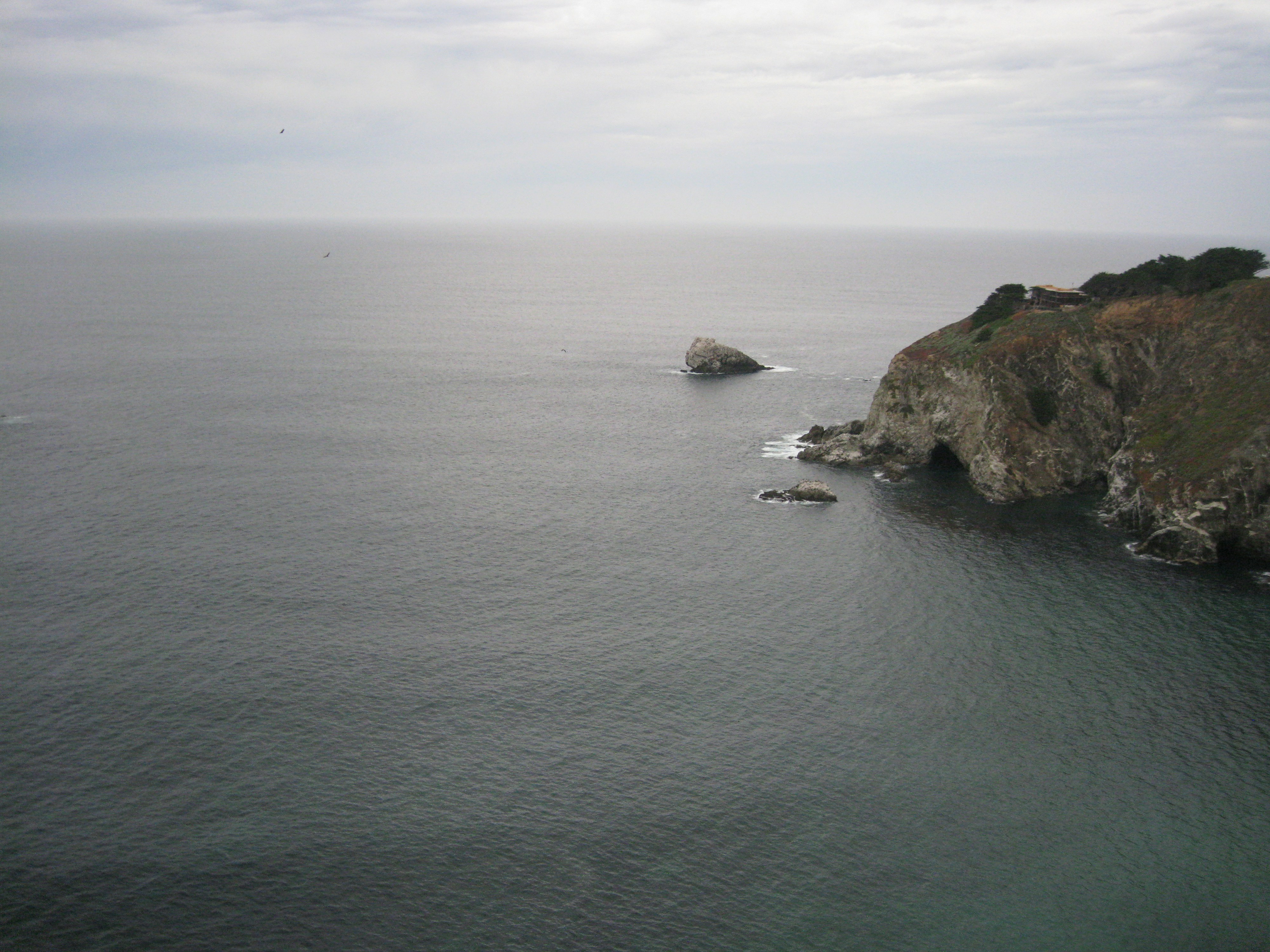 Coastal scene with a rugged cliff on the right meeting a calm, expansive sea under a soft, cloudy sky. The composition highlights the shoreline jut and a distant rock outcrop with muted color and gentle light.