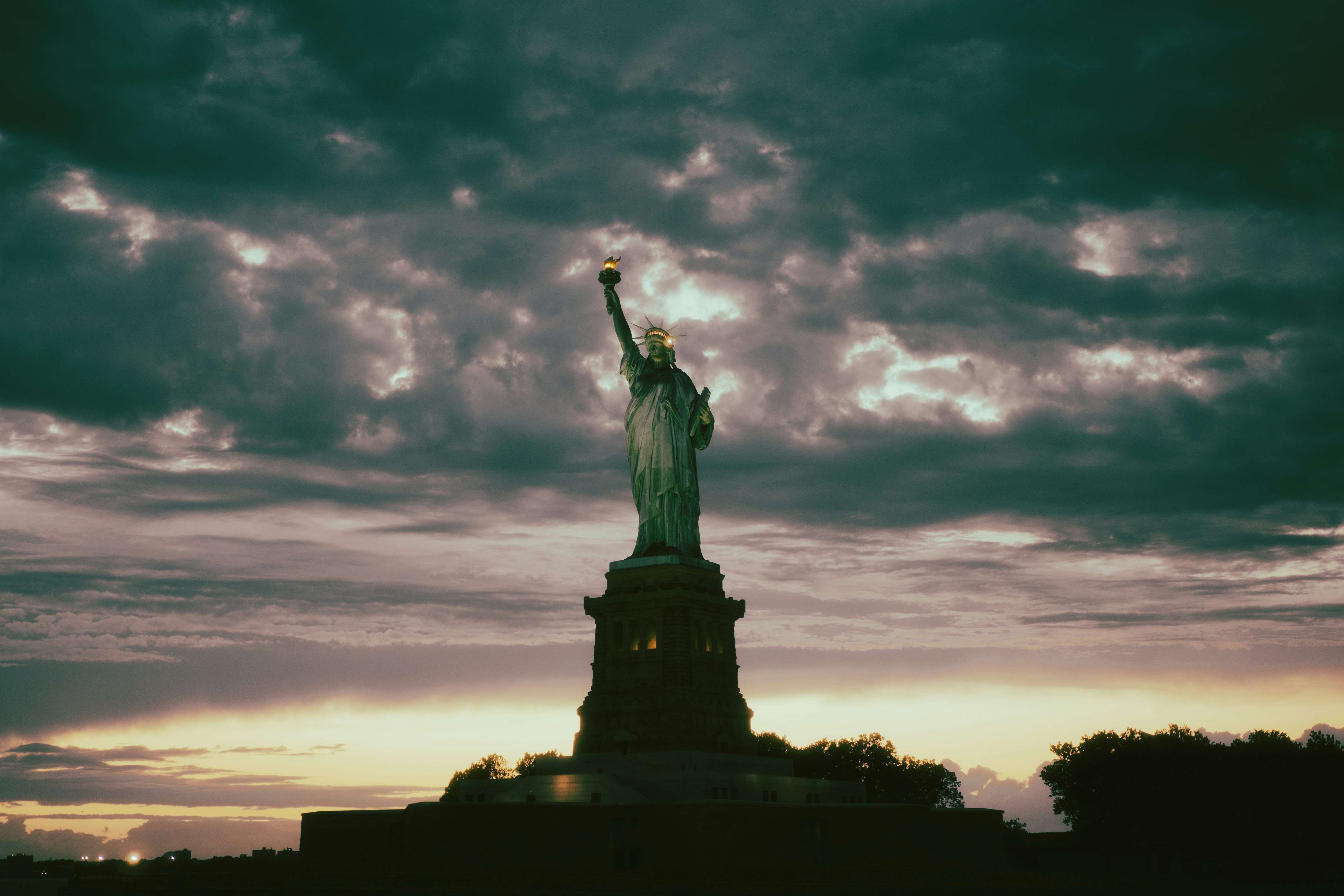 Silhouette of the Statue of Liberty rises above the dark base as a dramatic sunset sky unfolds, creating a moody, high-contrast scene.