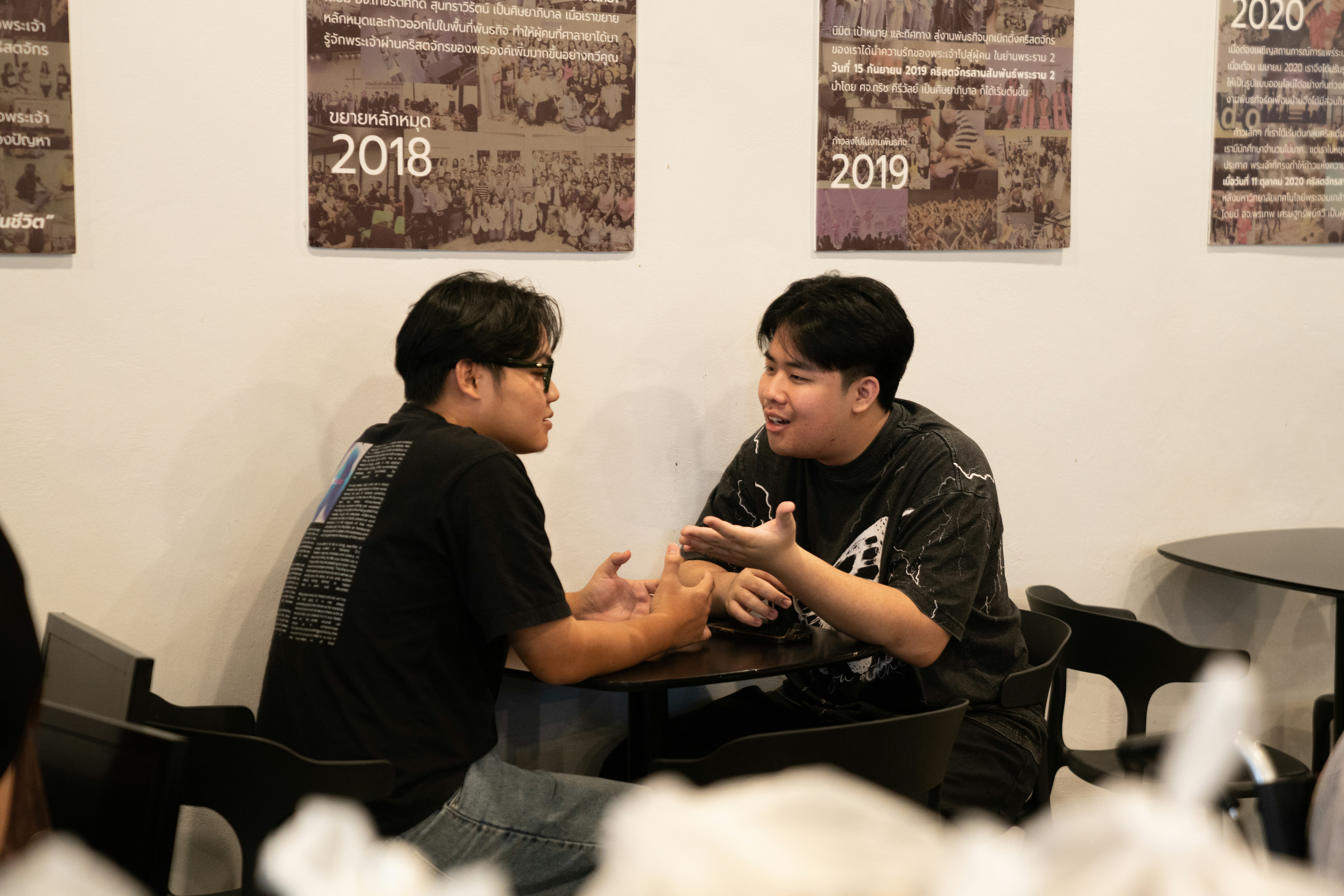 Two men sit across a small table in a casual café, engaged in conversation; posters line the wall behind them, creating a warm, social ambiance.