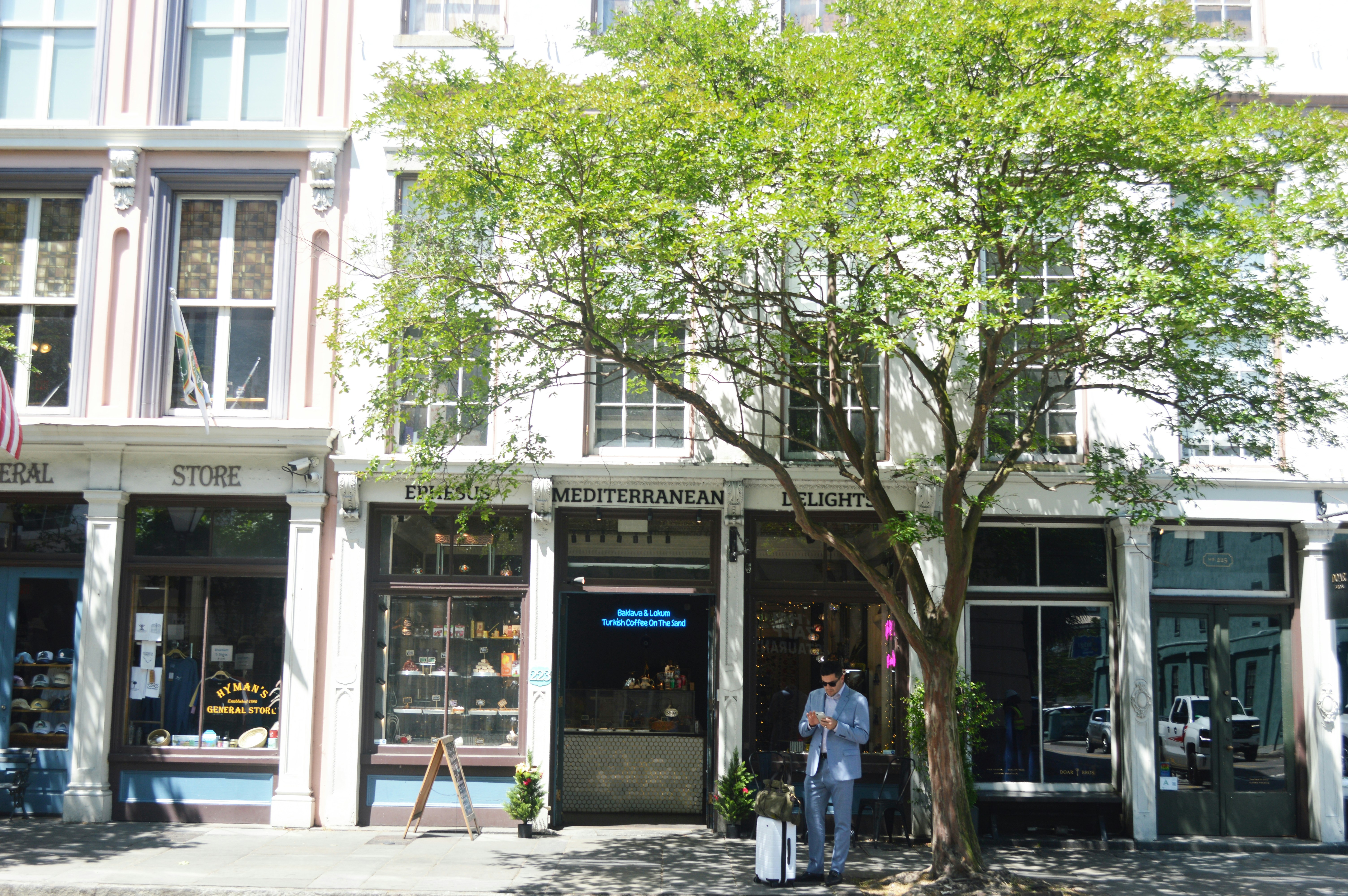 Sunlit storefronts and a traveler pausing beneath a leafy avenue