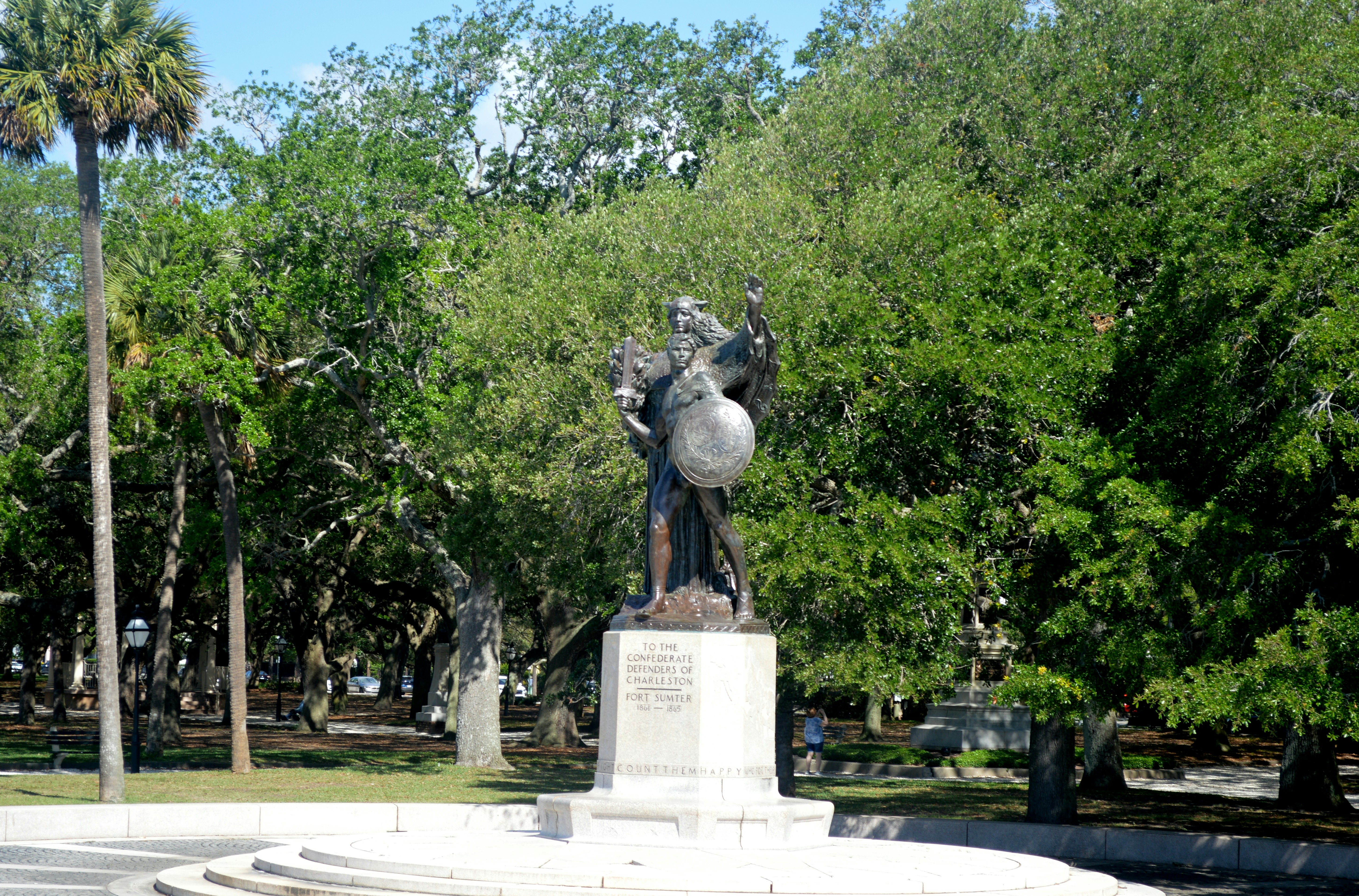 Statue atop a circular pedestal surrounded by a park with trees; sunlit scene emphasizing the sculpted figure with raised arms. A simple, outdoor monument photograph.