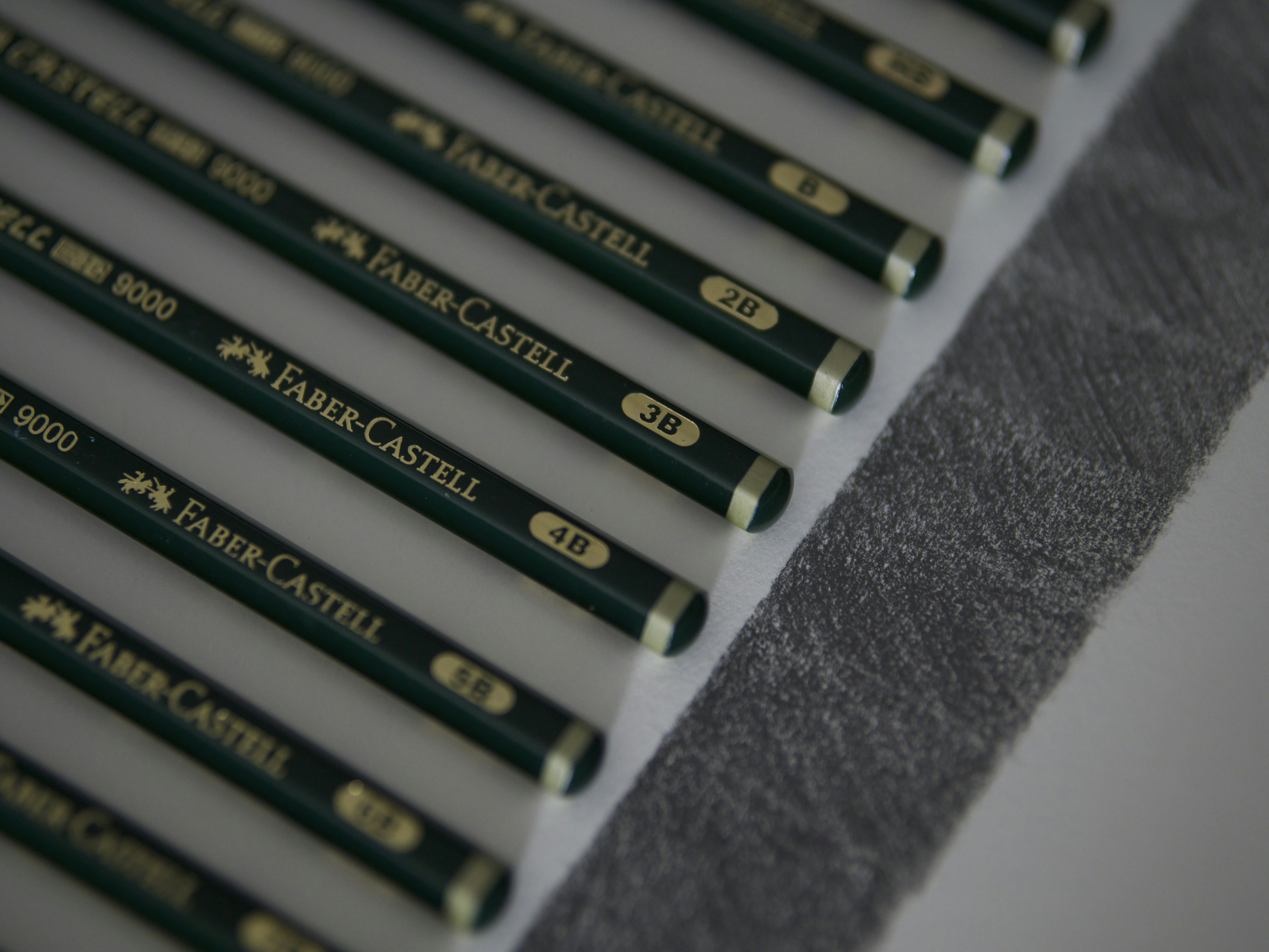 A close-up of green Faber-Castell pencils arranged diagonally across a textured surface, emphasizing the branded barrels and metallic ferrules. The composition highlights a linear study of color and texture.