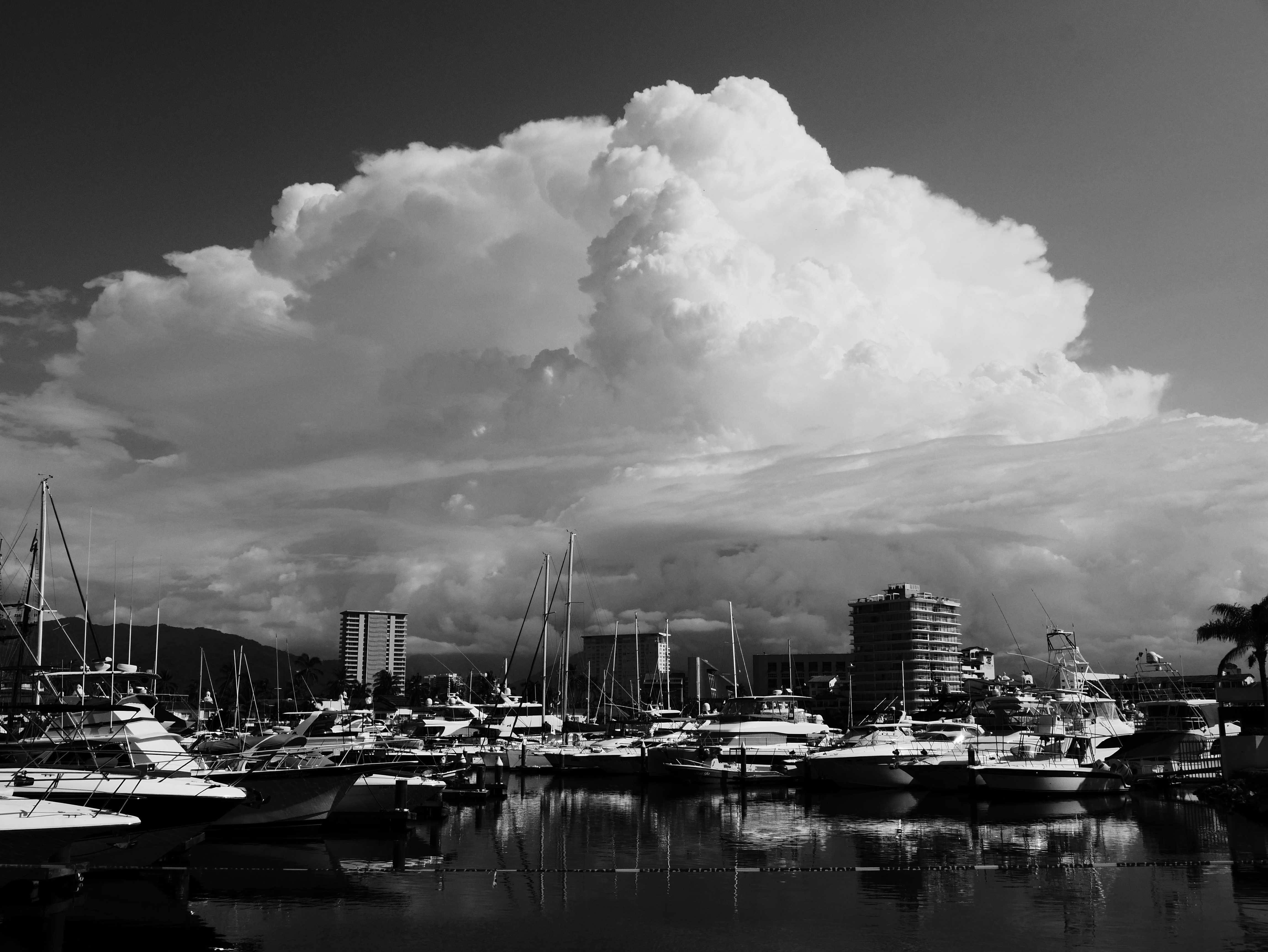 Monochrome harbor scene with moored boats and distant buildings reflected in calm water, emphasizing the dramatic sky.