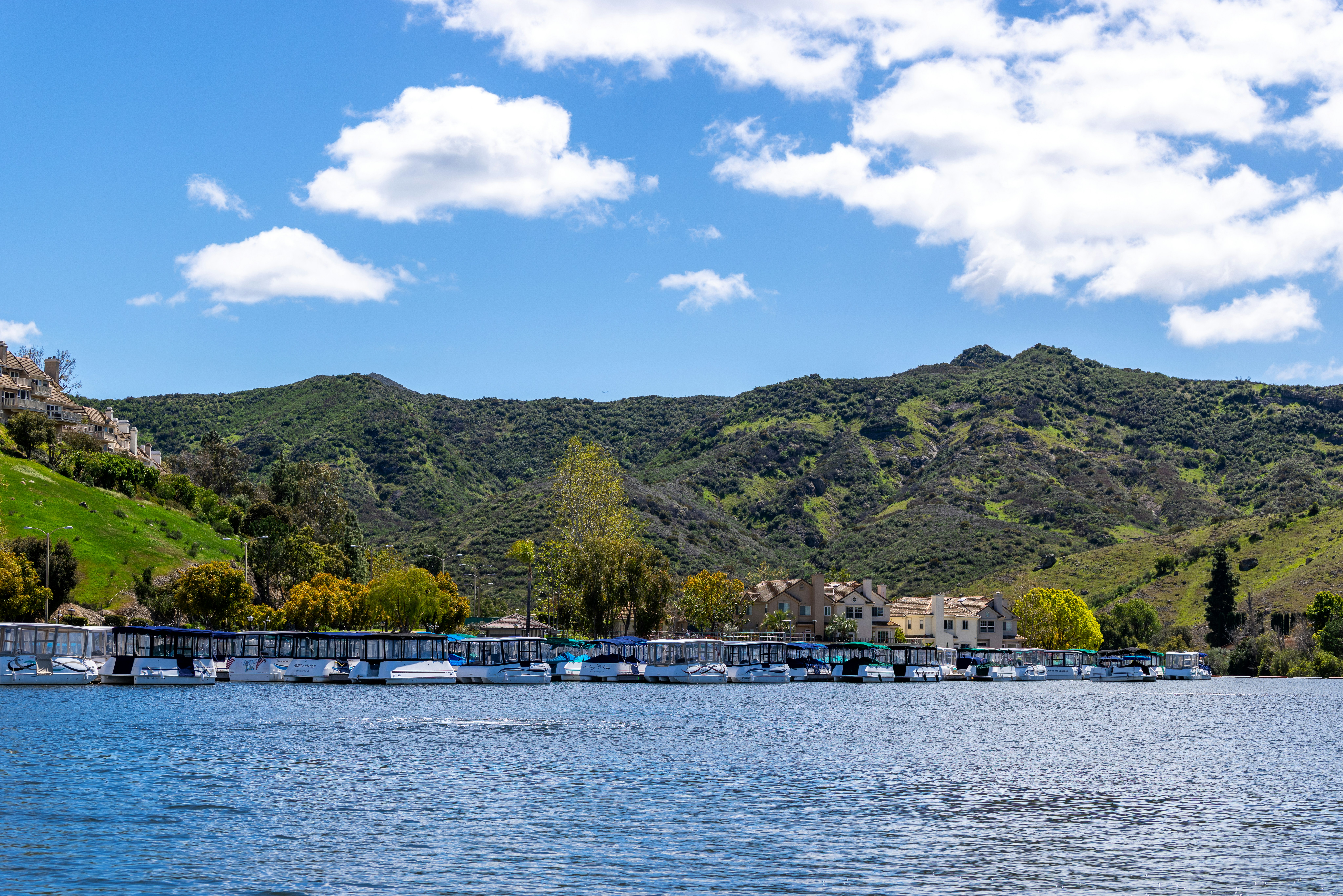 A tranquil lakeside scene with a row of buildings along the water and a backdrop of rolling green hills under a bright blue sky.
