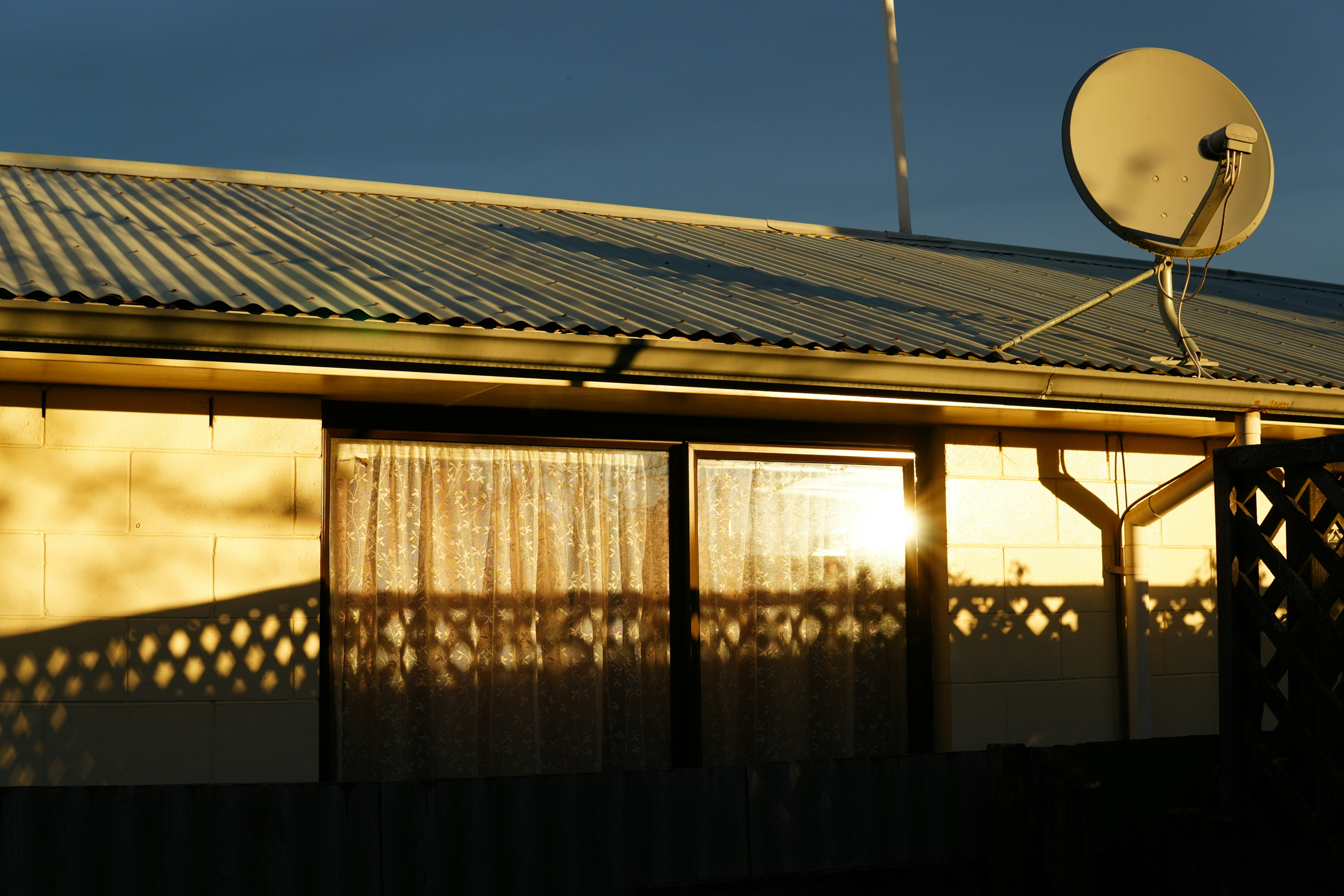 Sunlit roof with corrugated metal, satellite dish, and lace-curtained windows casting long shadows across the facade.