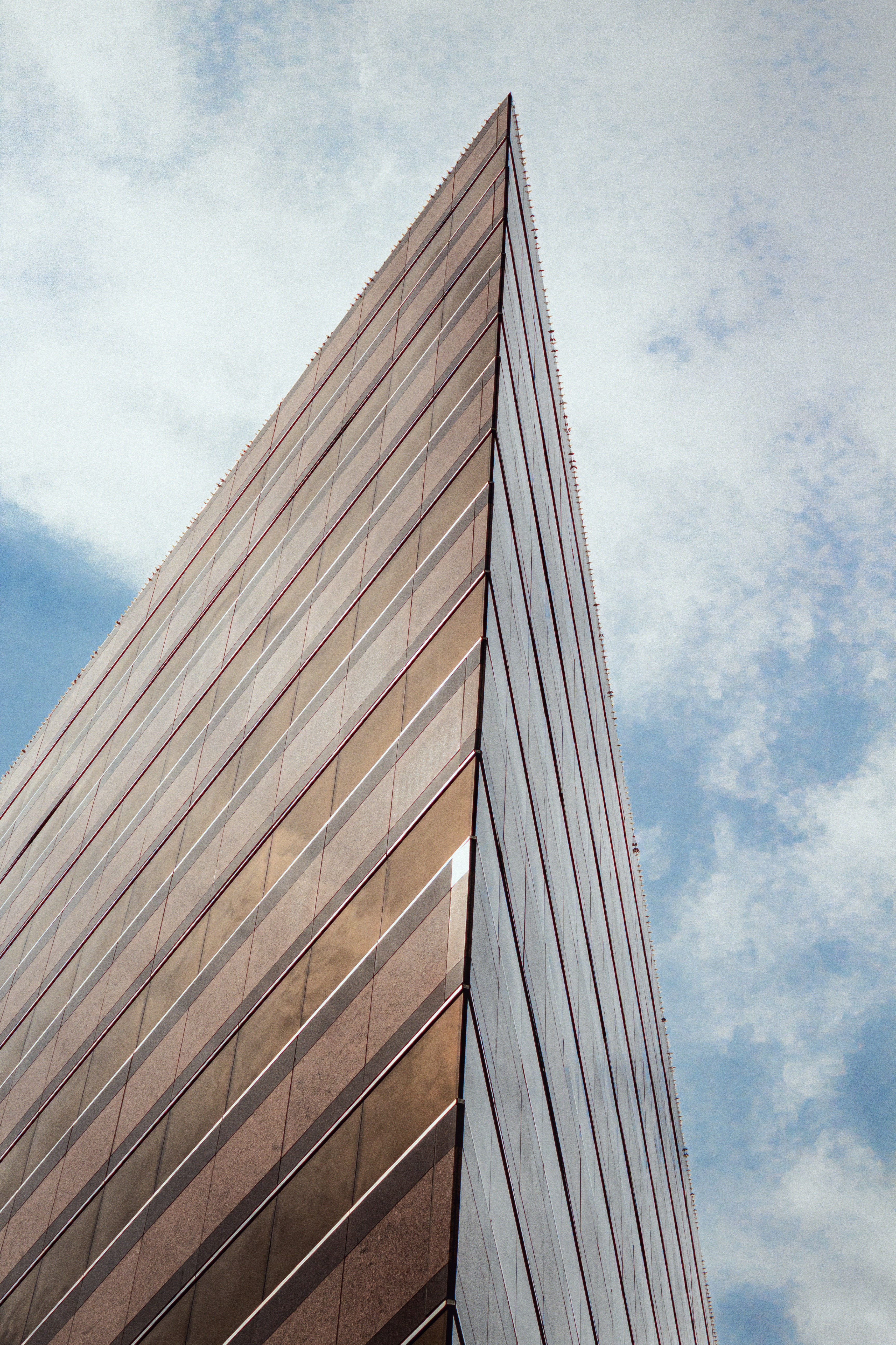 Sleek glass-clad skyscraper angled toward the sky against a blue, cloudy backdrop. A tall, reflective surface dominates the frame, emphasizing geometry and height.