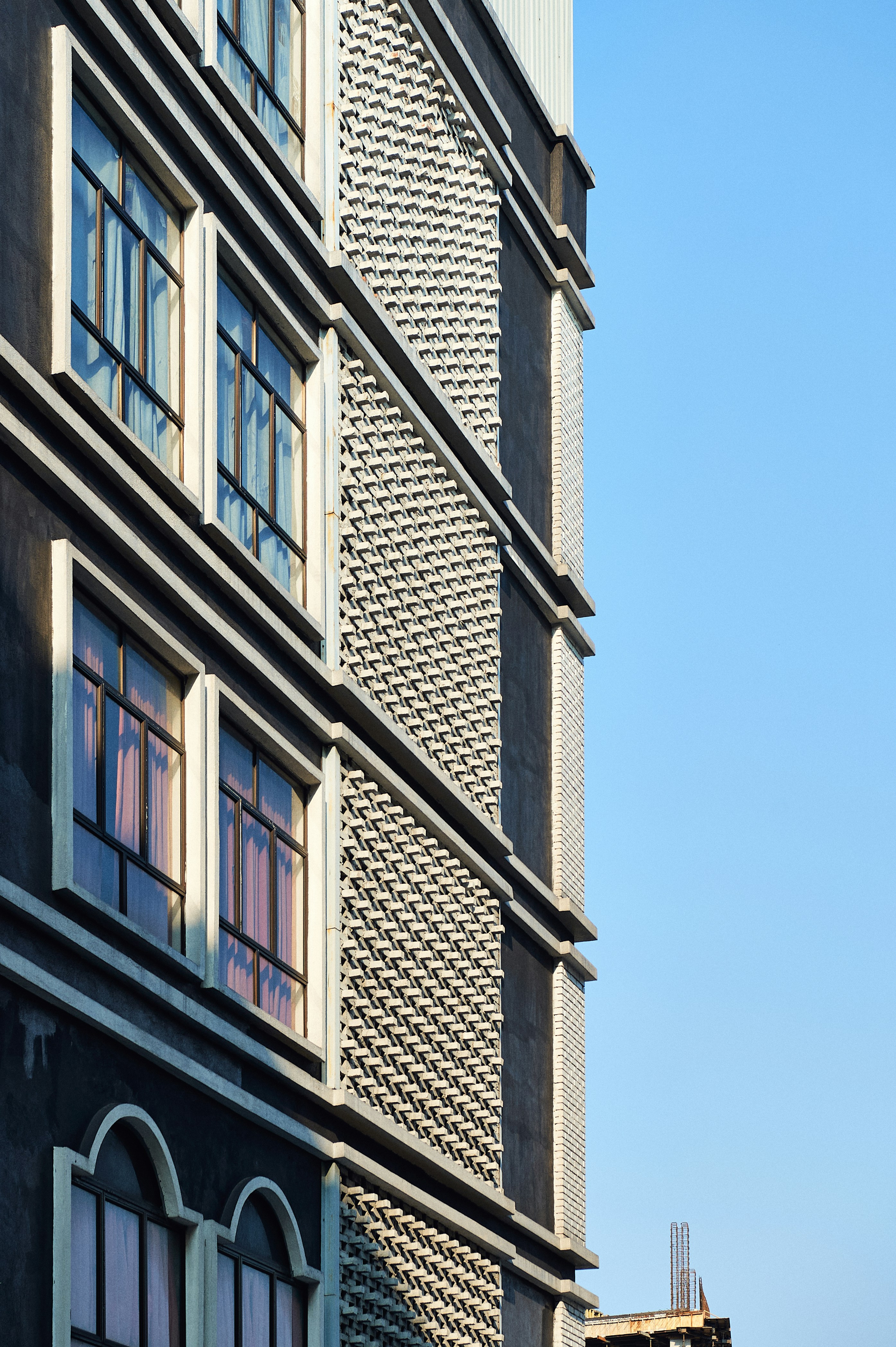 Tall city facade with geometric brick pattern and window bays bathed in clear daylight against a blue sky. Urban architecture detail with repeating textures and subtle shadows.