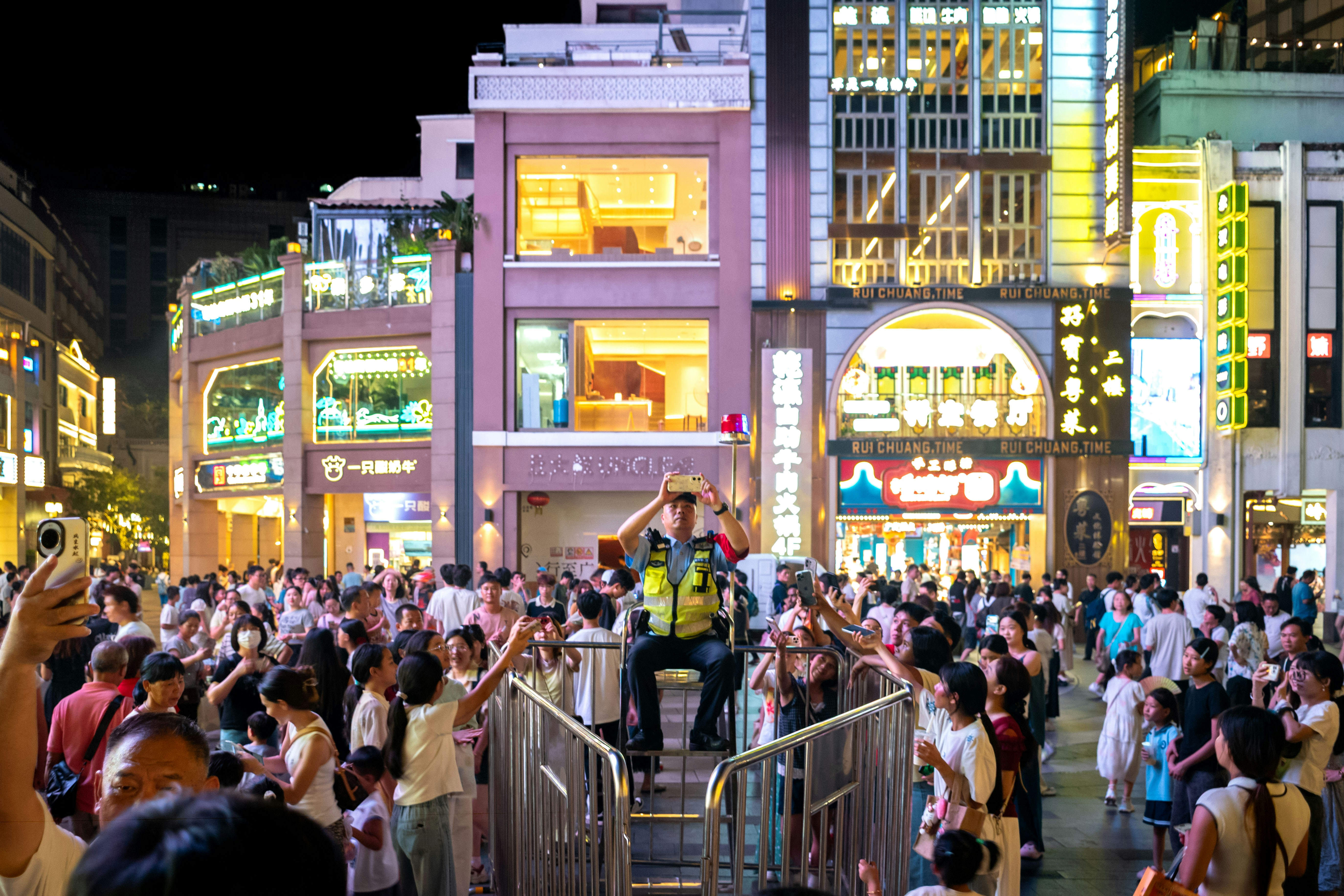 Crowd-filled night scene at a bustling urban plaza with a performer on a raised platform under bright storefronts and neon signs.
