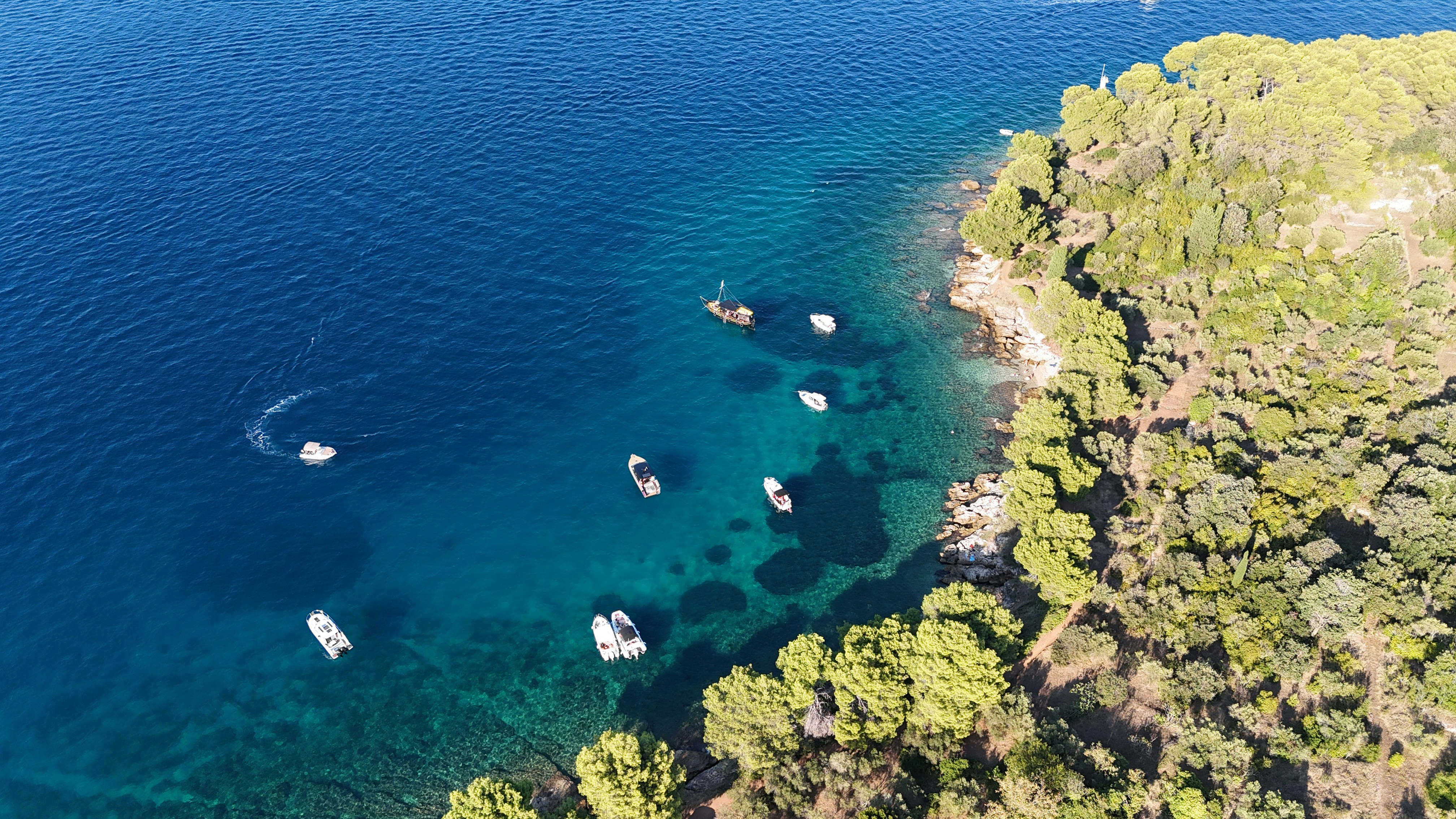 Turquoise coastal water with several anchored boats near rocky shoreline and pine-covered cliffs, captured from an aerial view.