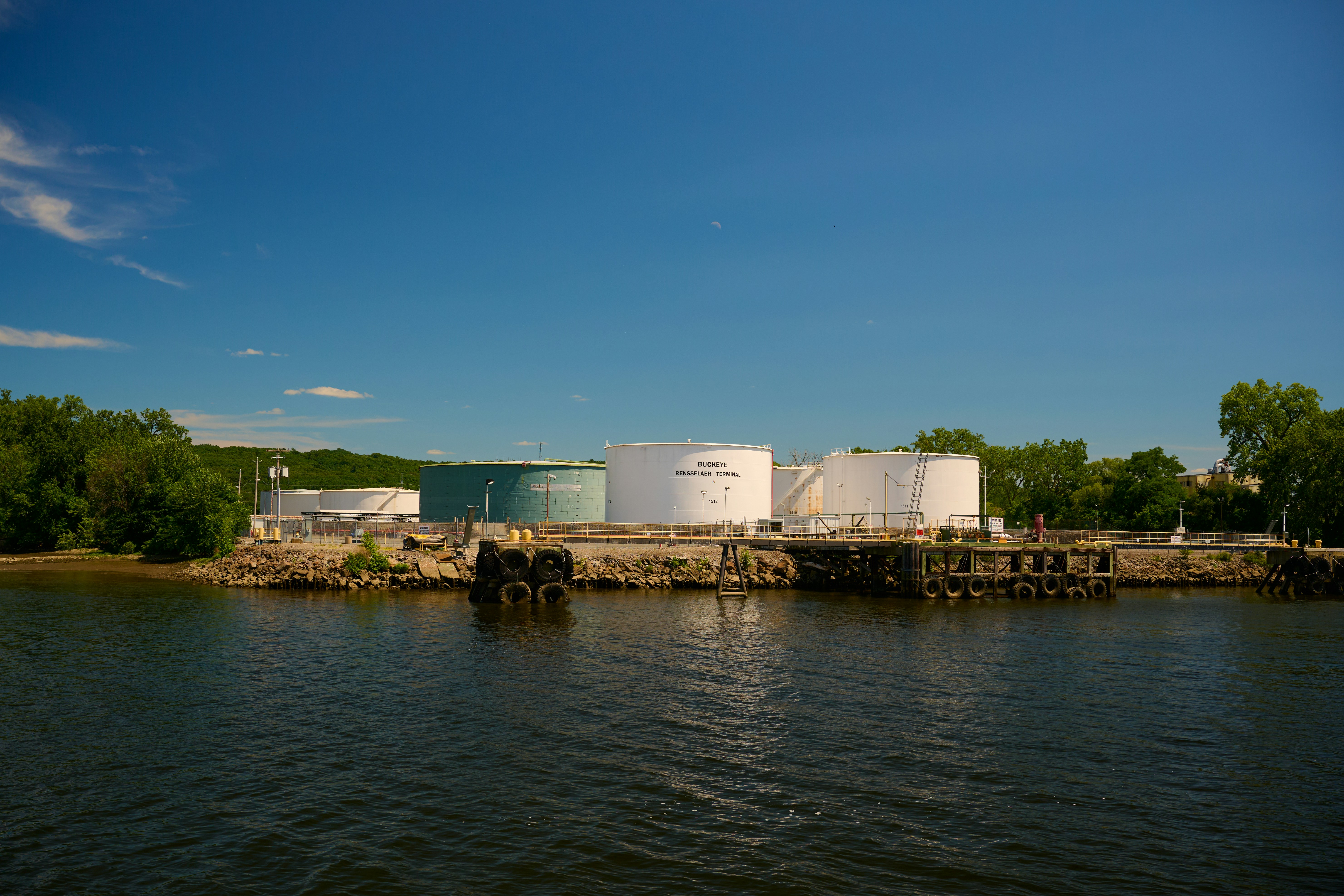Bright, calm waterfront view of a refinery or storage tanks along a pier with trees in the background and a deep blue sky. The scene is unembellished and documentary, with a focus on the industrial shoreline.