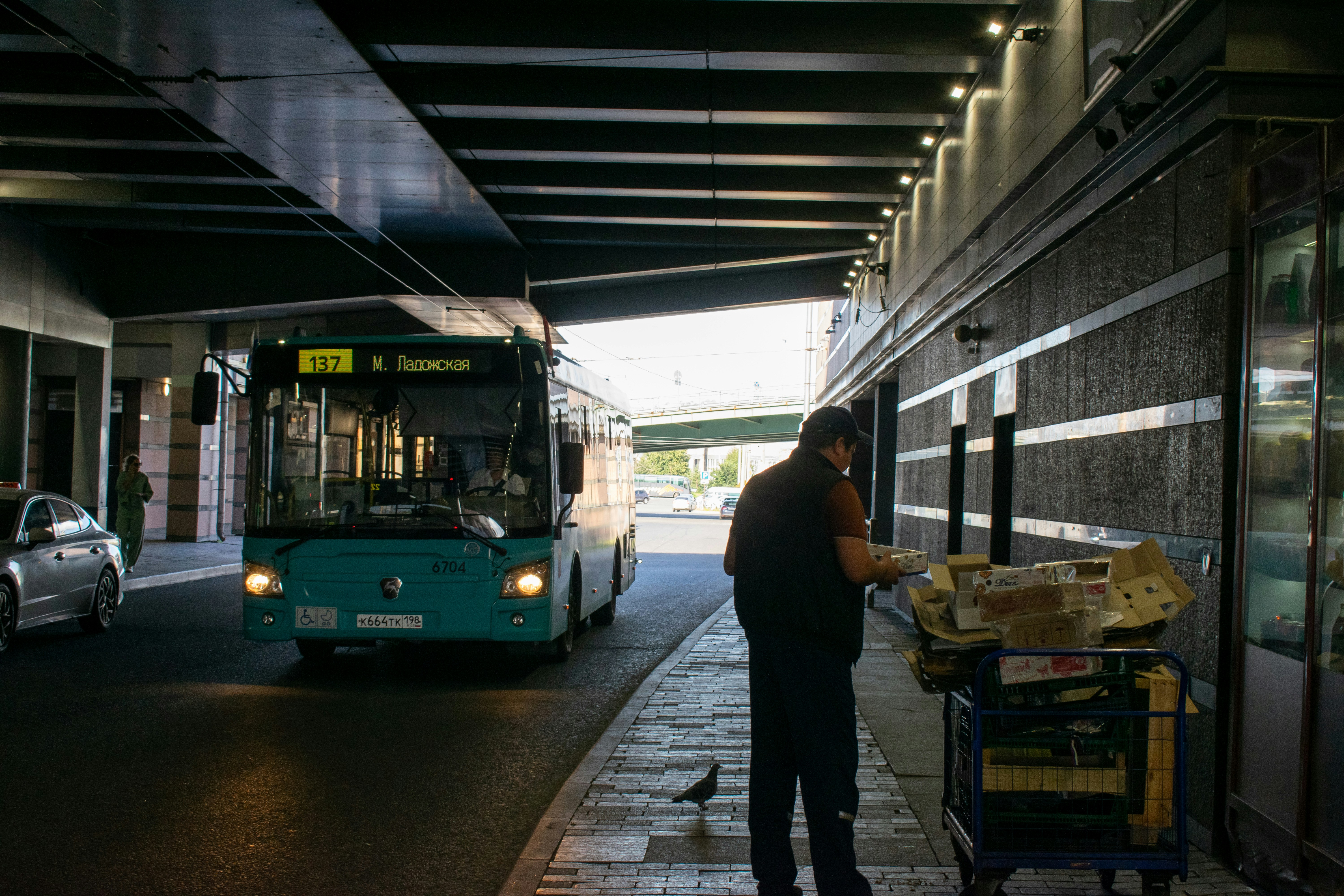 A city street scene under a covered walkway shows a turquoise bus approaching as a man sorts boxes at a vendor cart; a lone bird pecks nearby, with a worker and urban storefronts in the background.