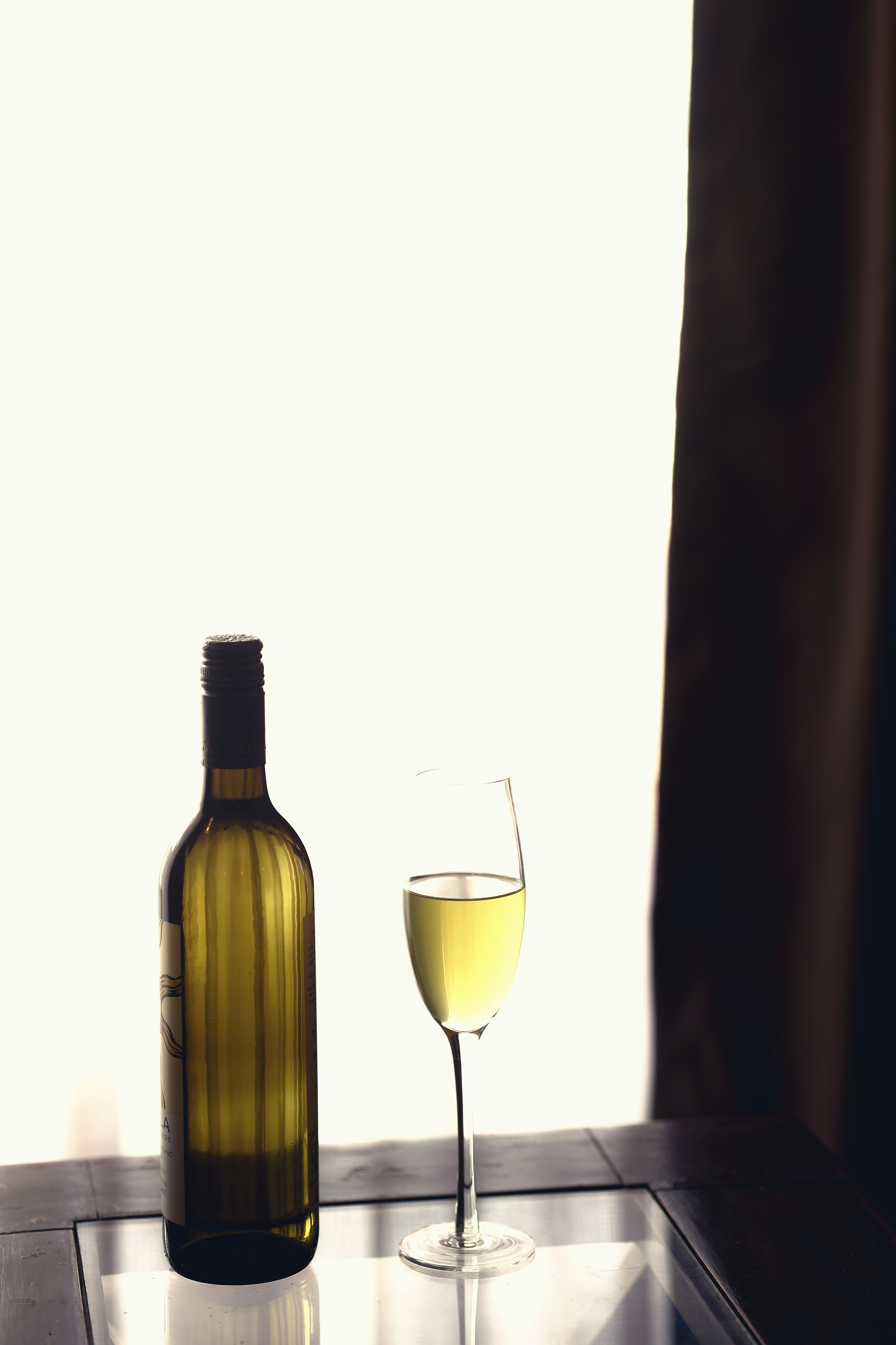 Backlit bottle and glass of white wine on a rustic table, with soft overexposure behind creating a warm, minimalist still-life.