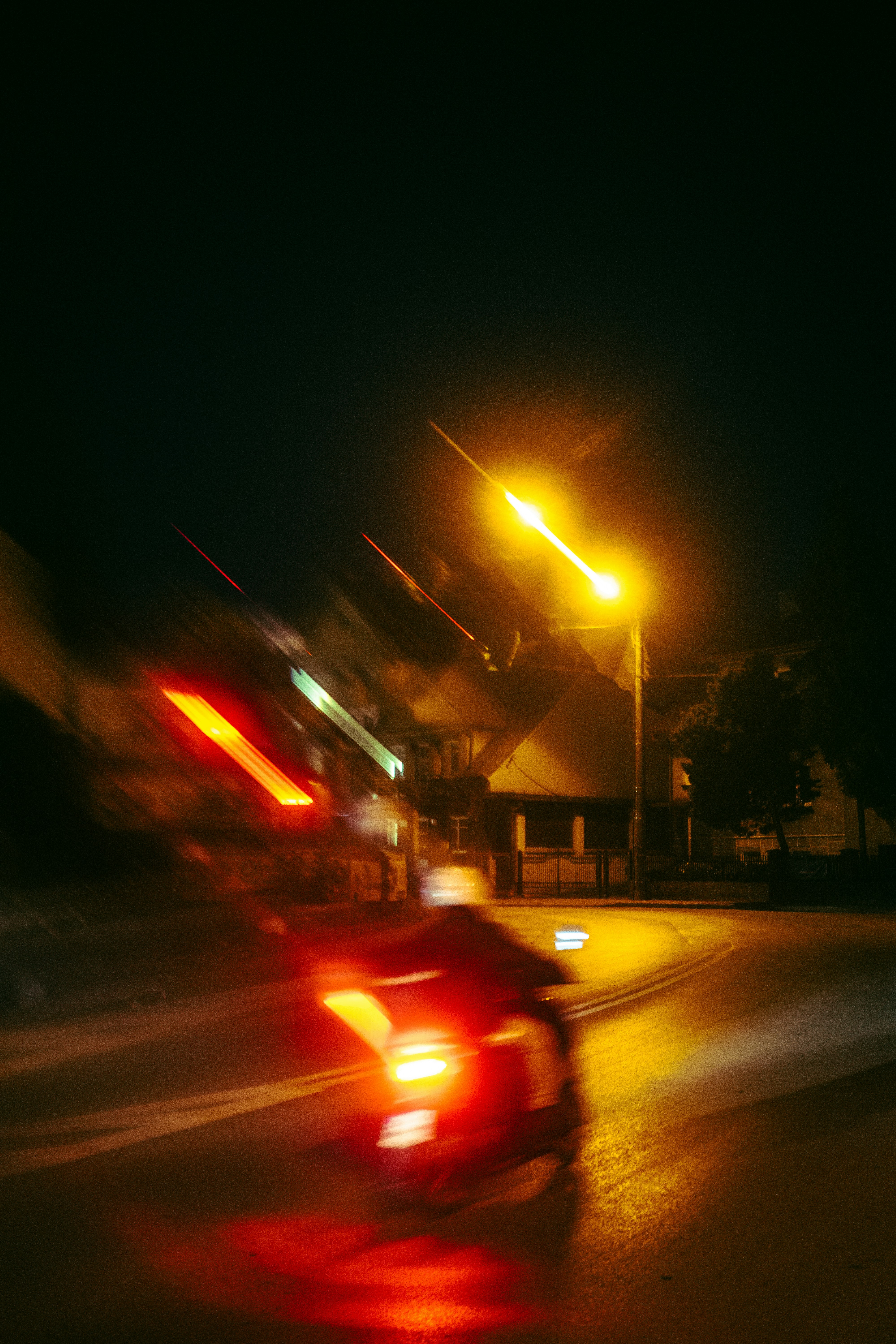 Motorbike rider glides past glowing street lamps in a dim, street-lit neighborhood, with motion blur giving a sense of speed and nighttime ambiance.