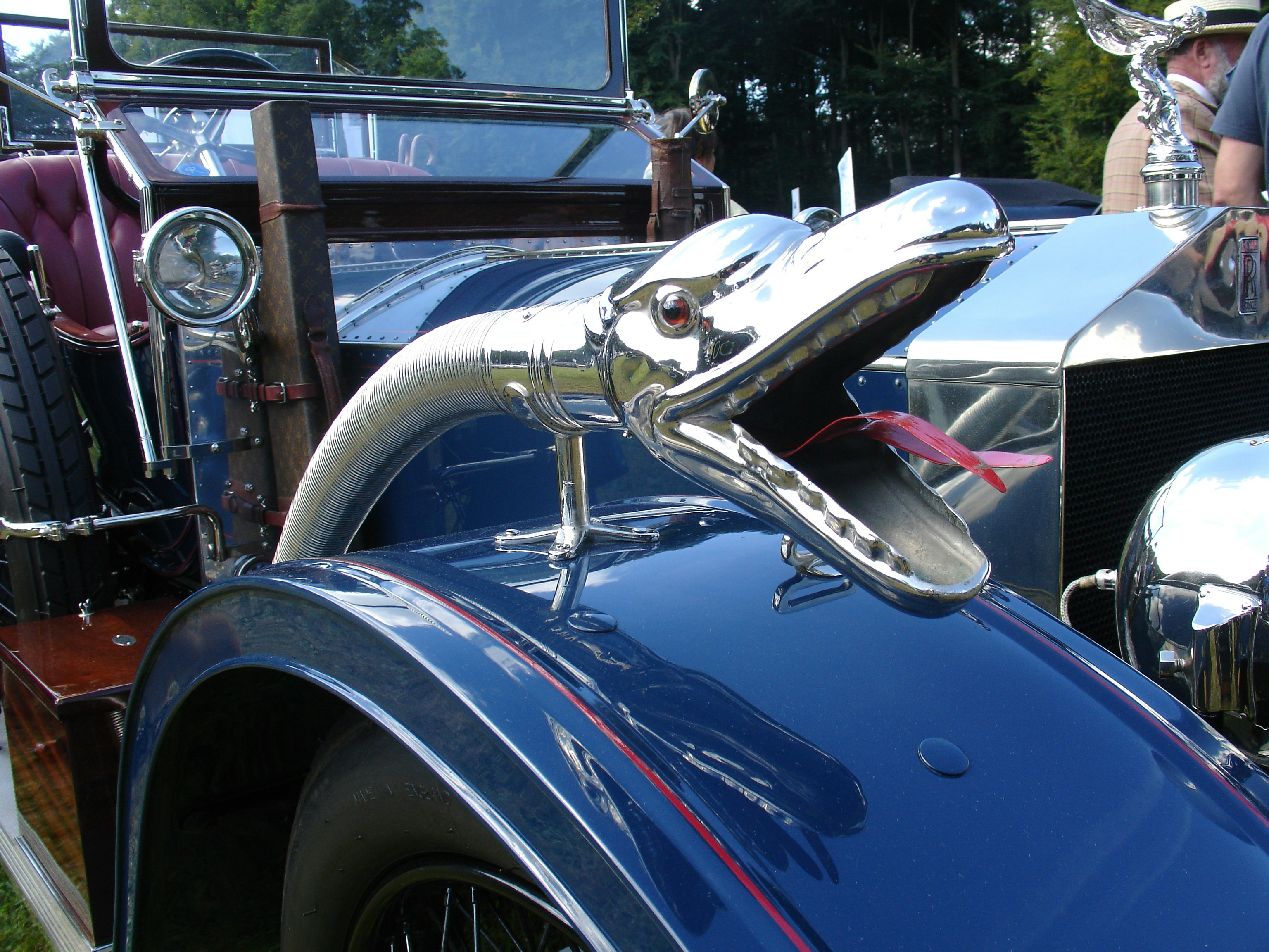 Blue vintage car with a chrome dragon hood ornament as the focal point, surrounded by other classic automotive details. A concise automotive still emphasizing the sculpture and metallic reflections.