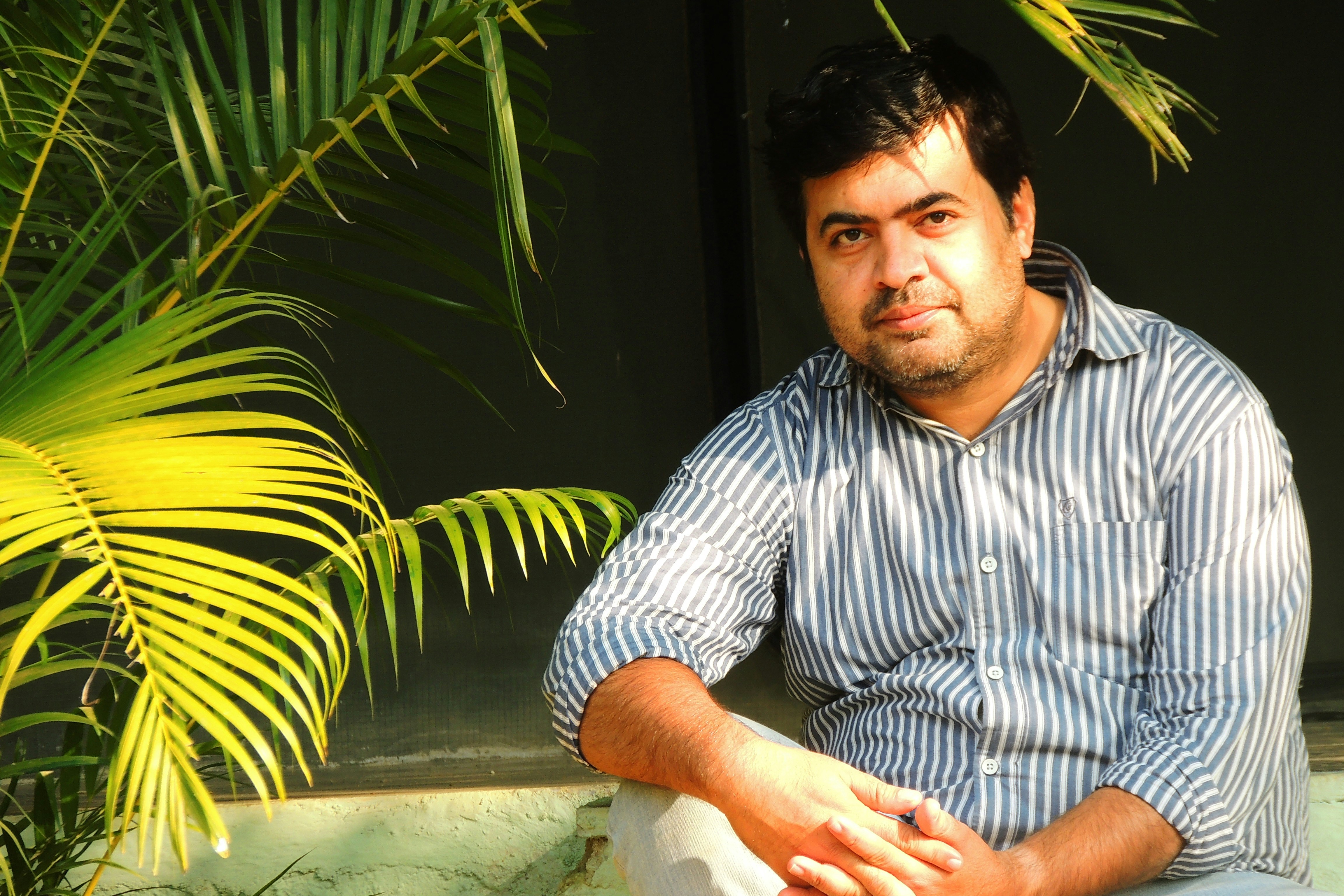 Portrait of a man in a striped shirt seated near tropical foliage with a dark background. A calm, outdoor-style composition emphasizing the subject’s face and upper body.