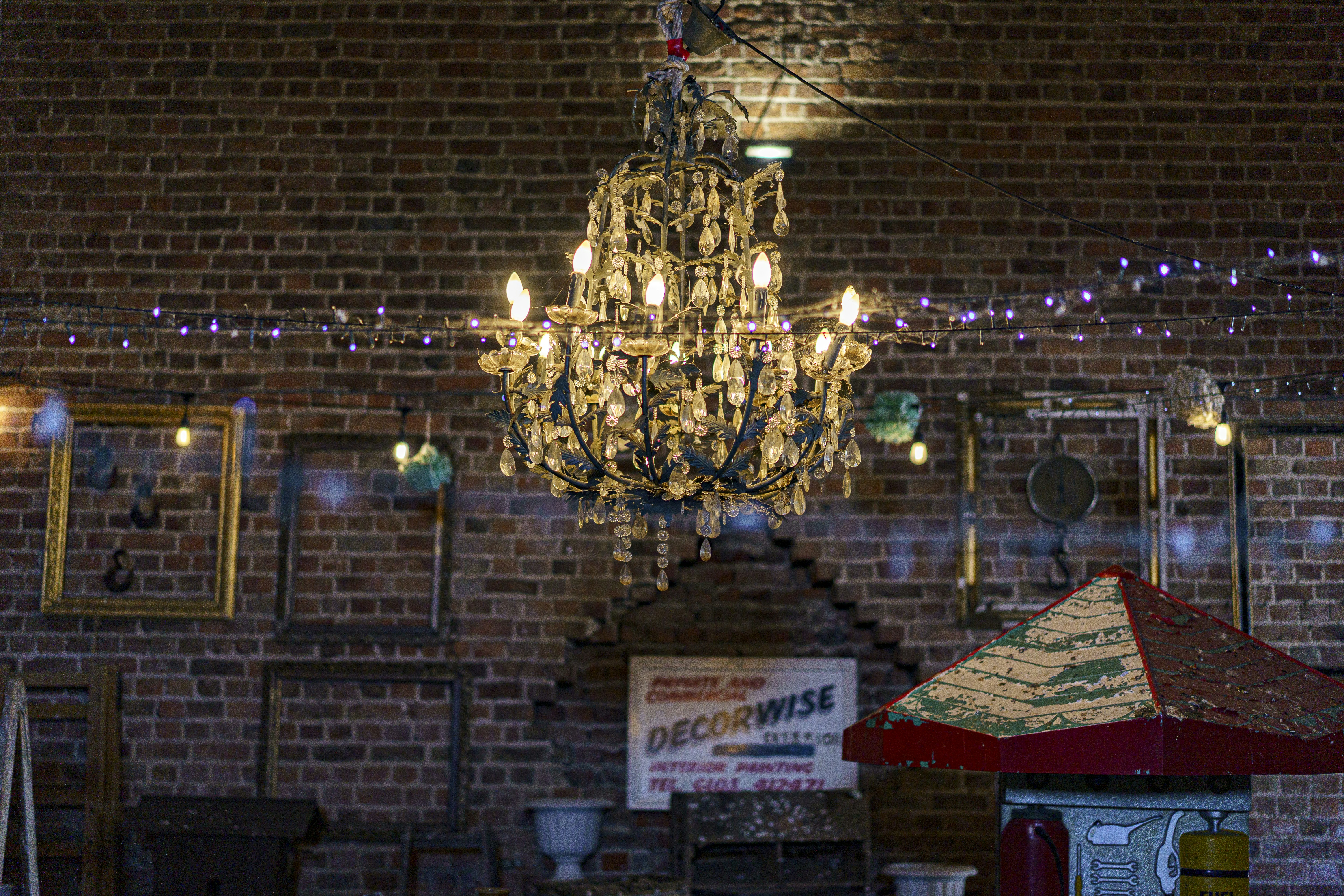 Chandelier hanging against a brick wall with string lights and a vintage sign in the background.